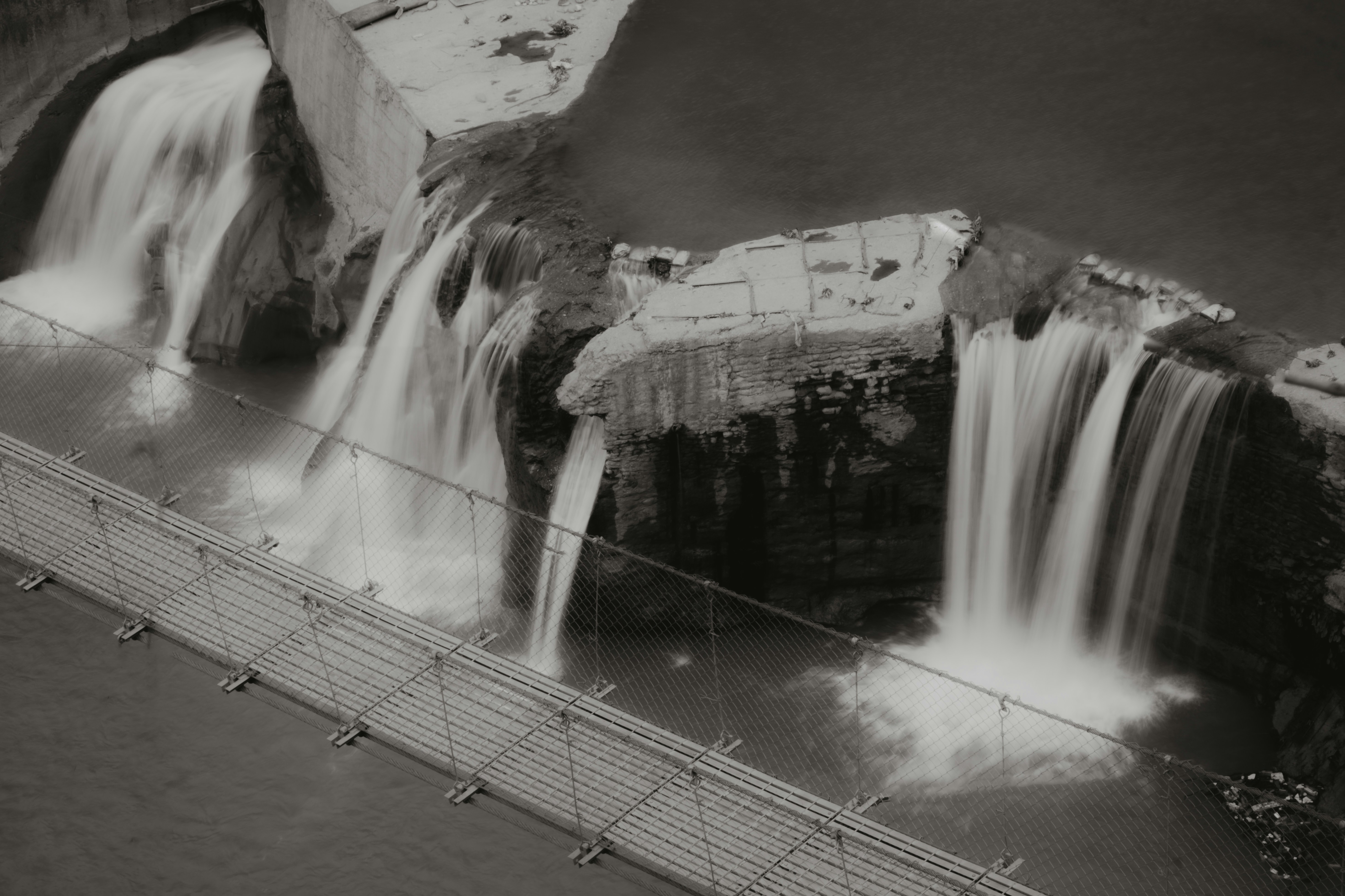 Monochrome Cascade: Water Unfolding Over the Dam Framed by a Chain-link Bridge