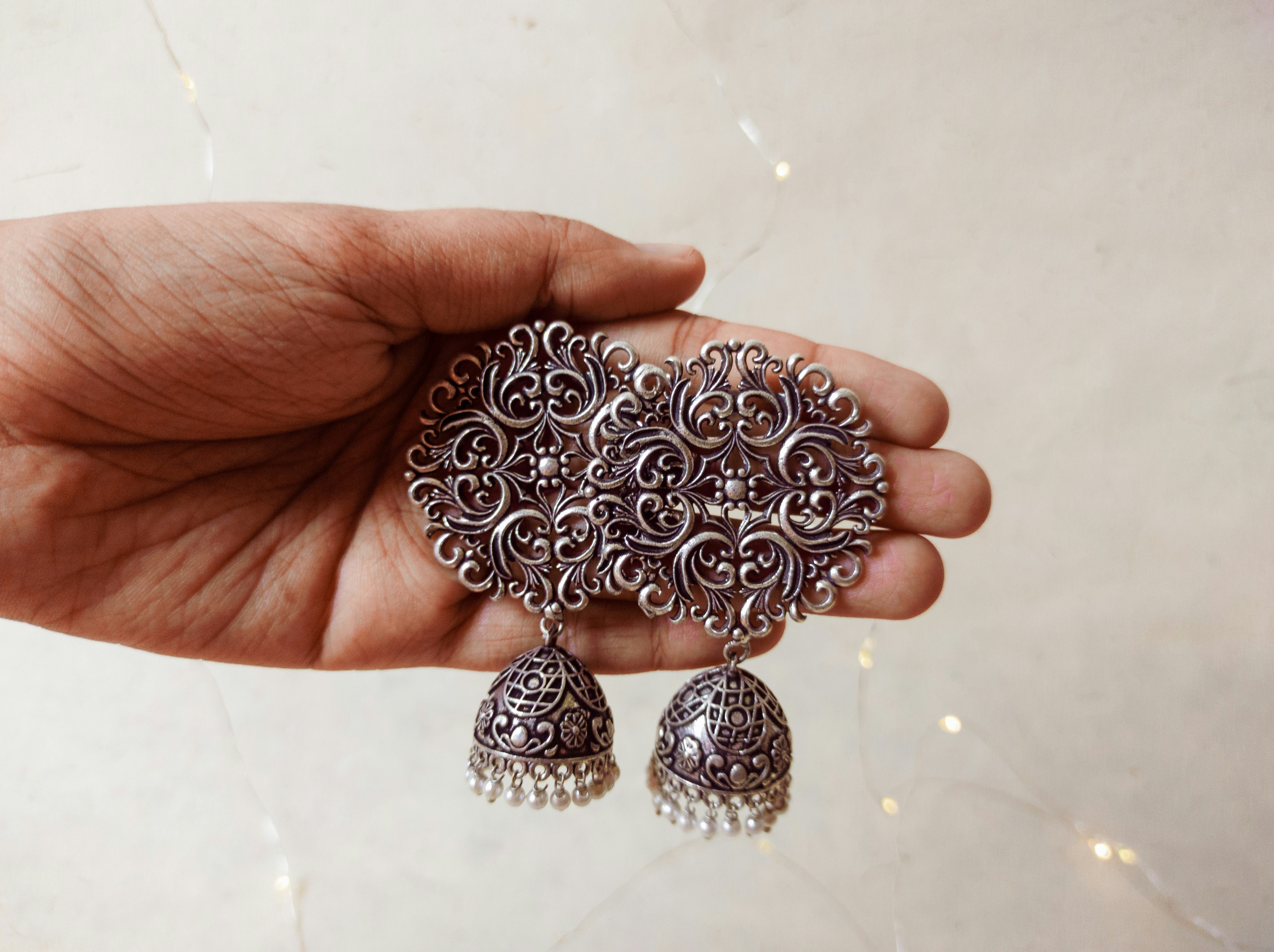 Close-up photograph of an ornate silver filigree jewelry piece with two dangling jhumka earrings, held by a hand against a softly lit background.