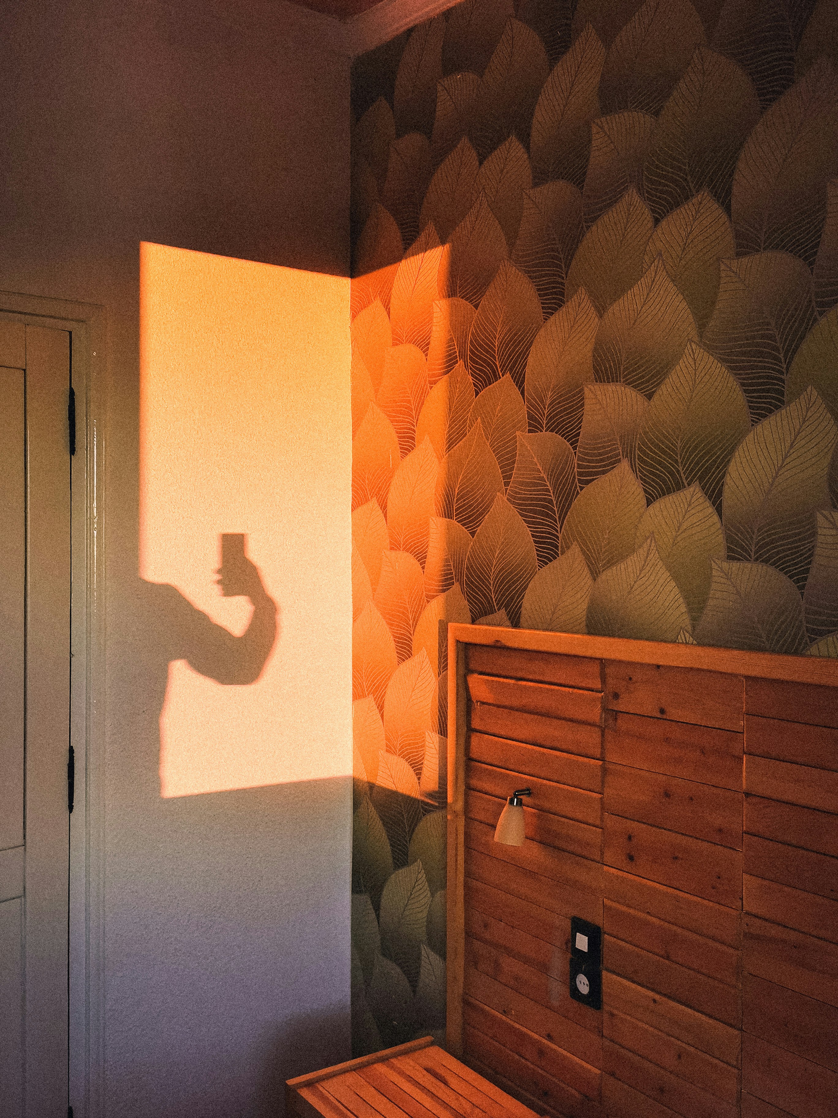 Shadow of a person holding a smartphone is projected onto a leaf-patterned wall, with a wooden headboard and warm orange light filling the corner.