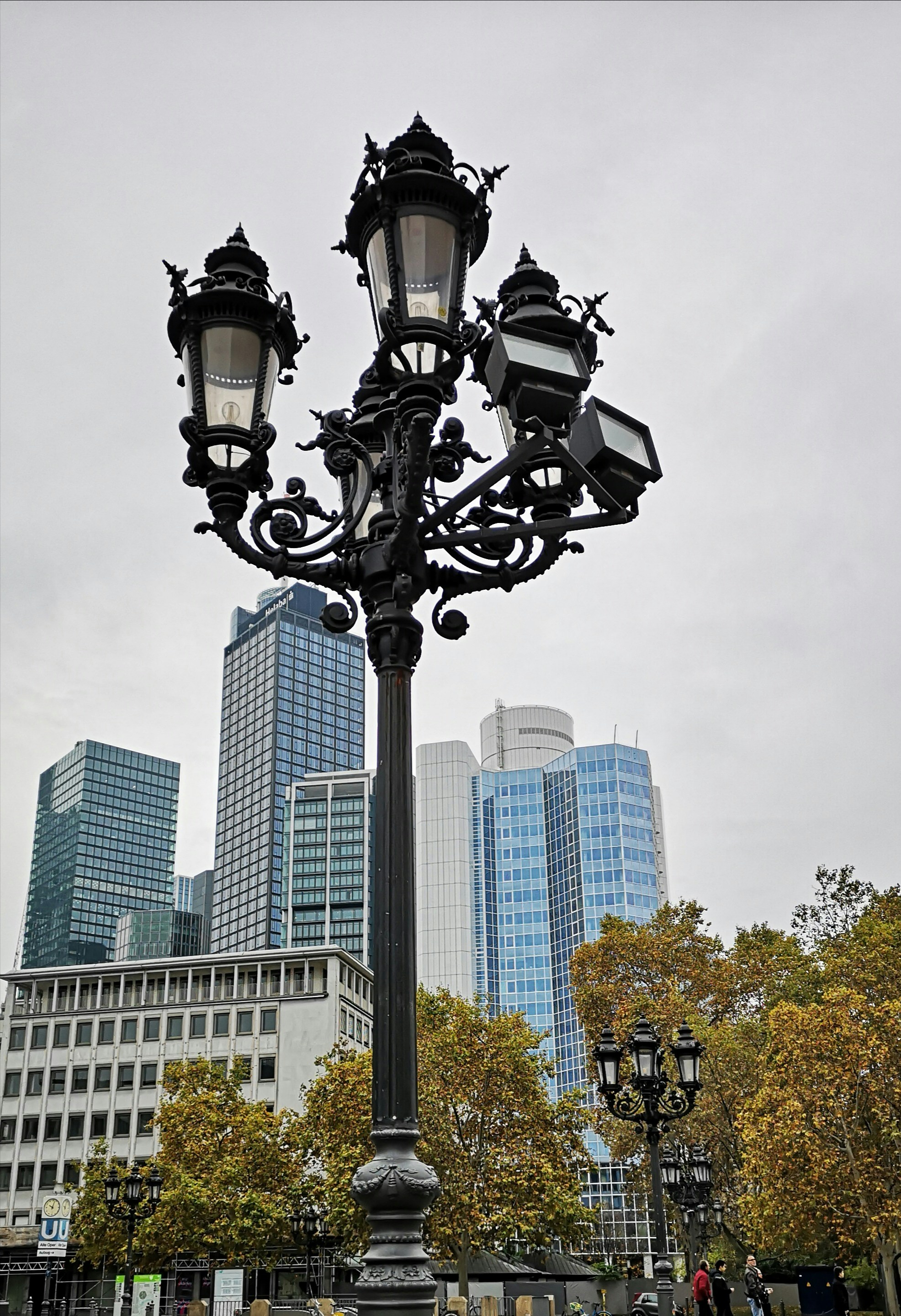 A decorative street lamp in the foreground against a backdrop of modern glass office buildings and autumn trees.