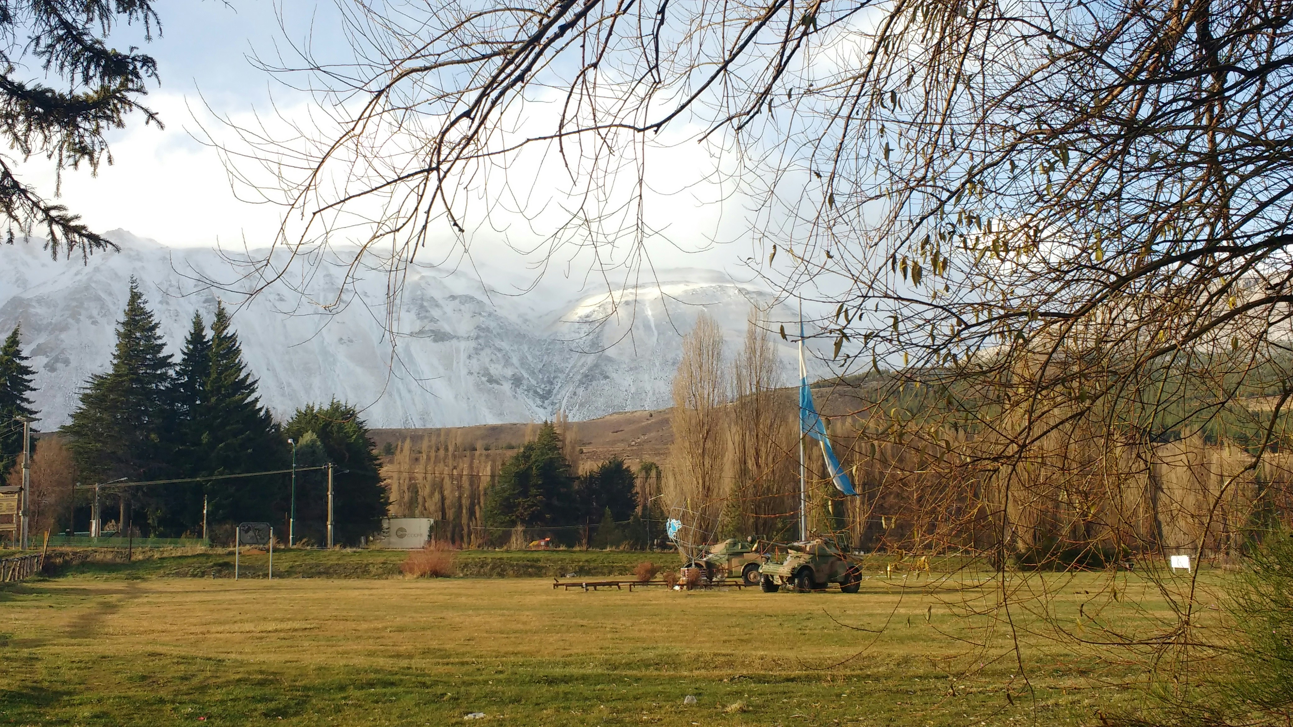 Wide landscape of a grassy field with a blue construction crane near center-right. Distant snow-dusted mountains loom under a clear sky.