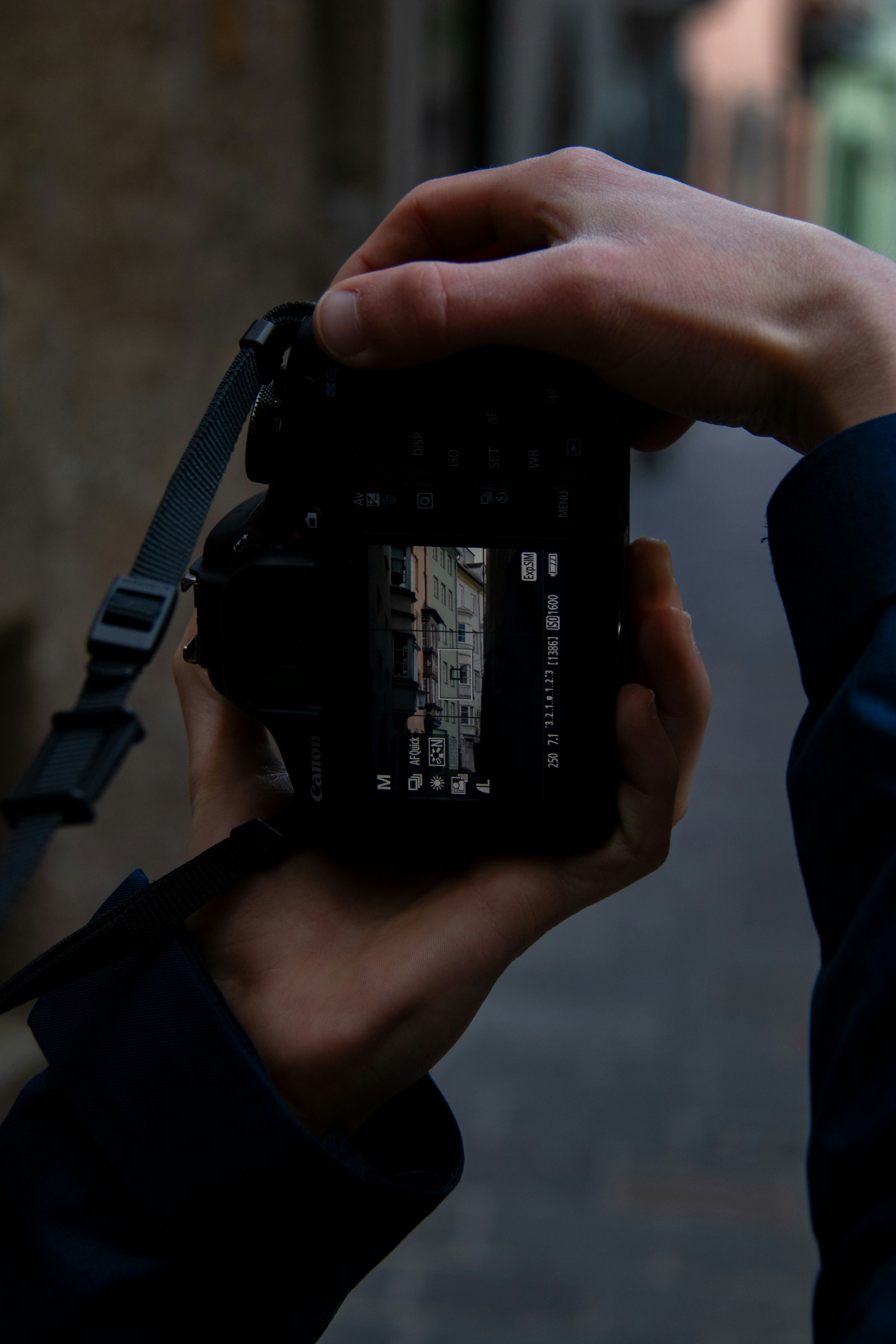 A photographer holds a Canon EOS 800D, capturing a narrow street scene reflected on the camera's LCD screen.