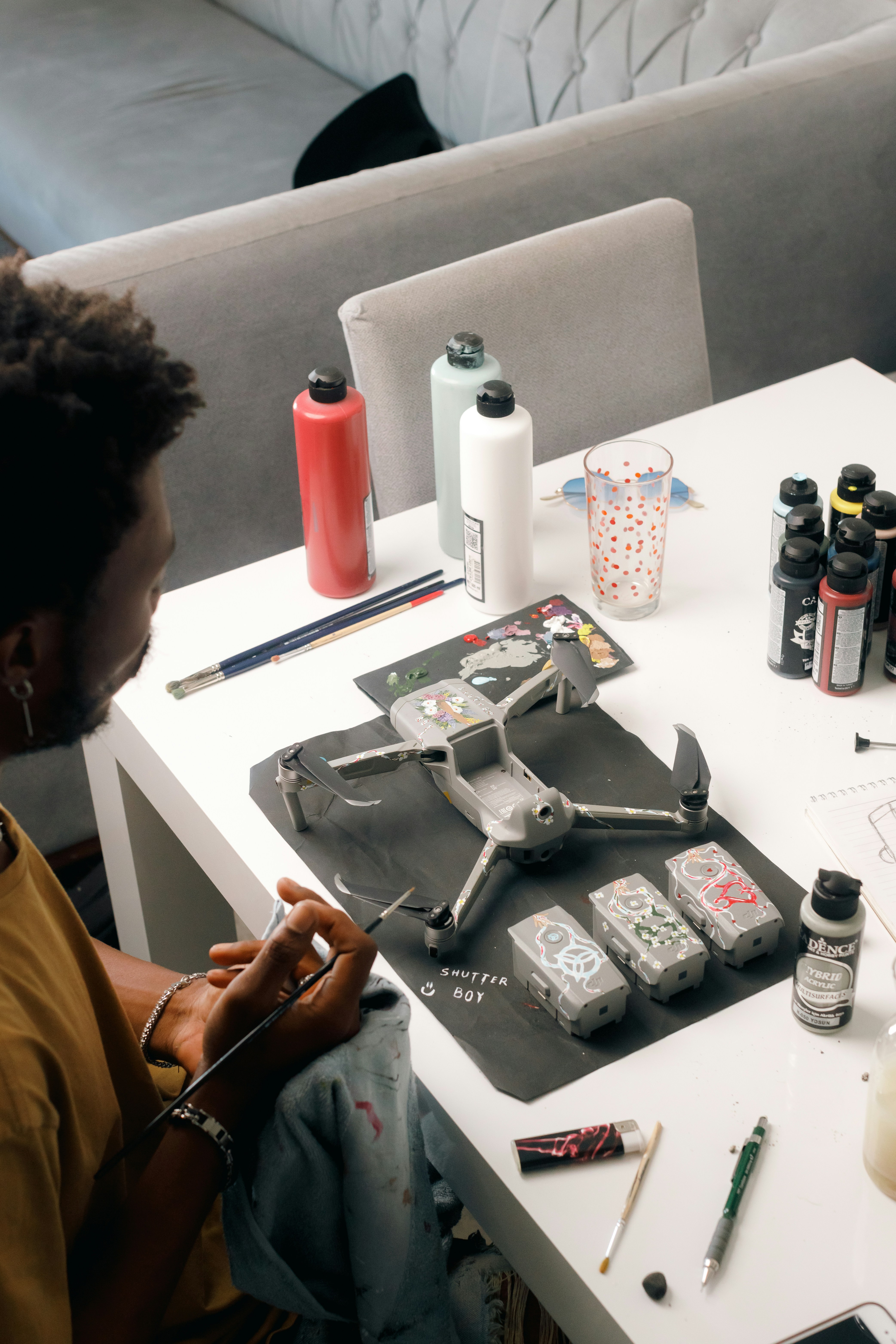 Artist sits at a white table with paints, brushes, and a small mechanical drone model, surrounded by markers and tools in a bright studio space.