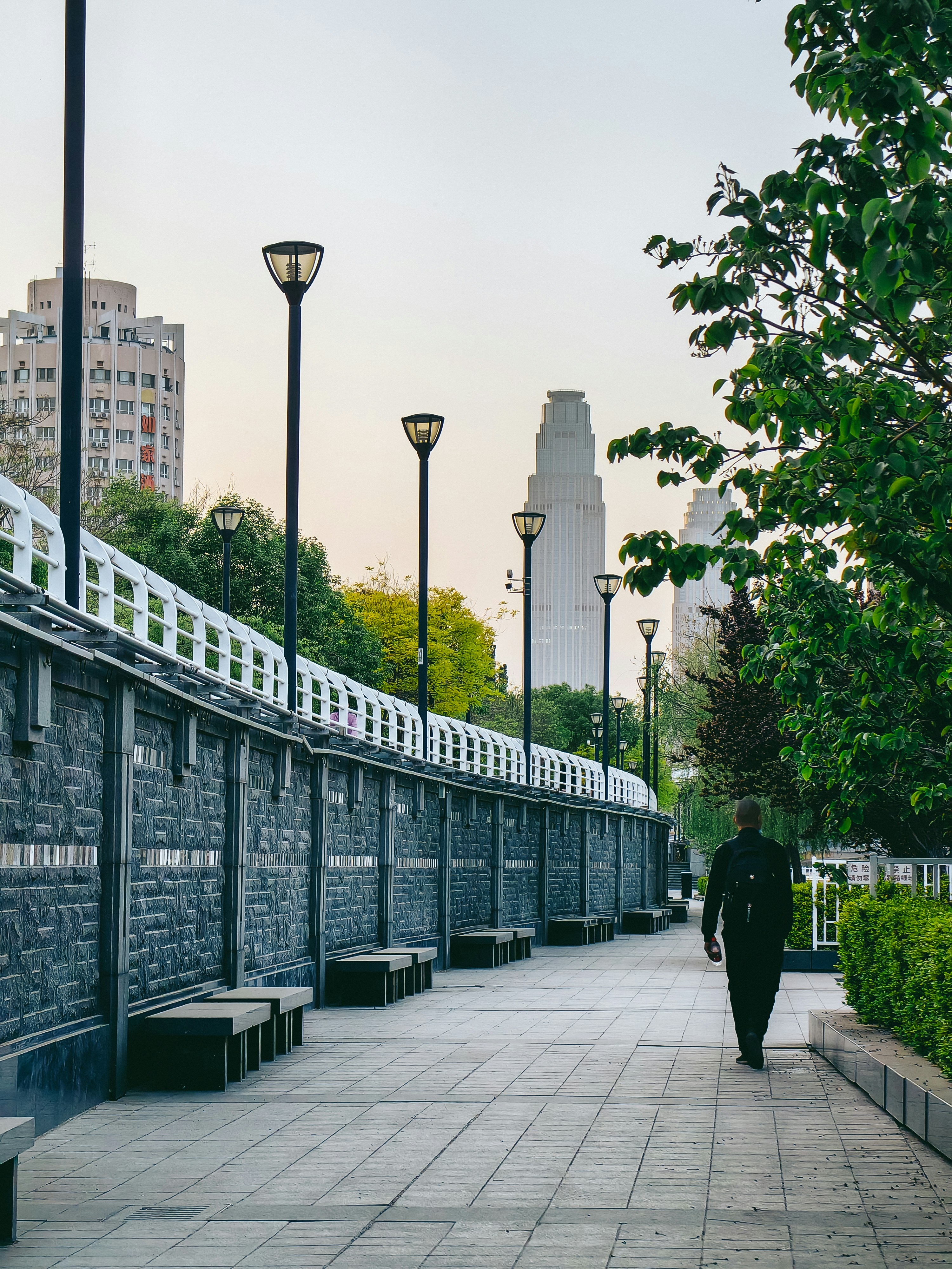 A lone figure walks along a paved promenade beside a brick wall, flanked by modern street lamps with a distant city skyline.