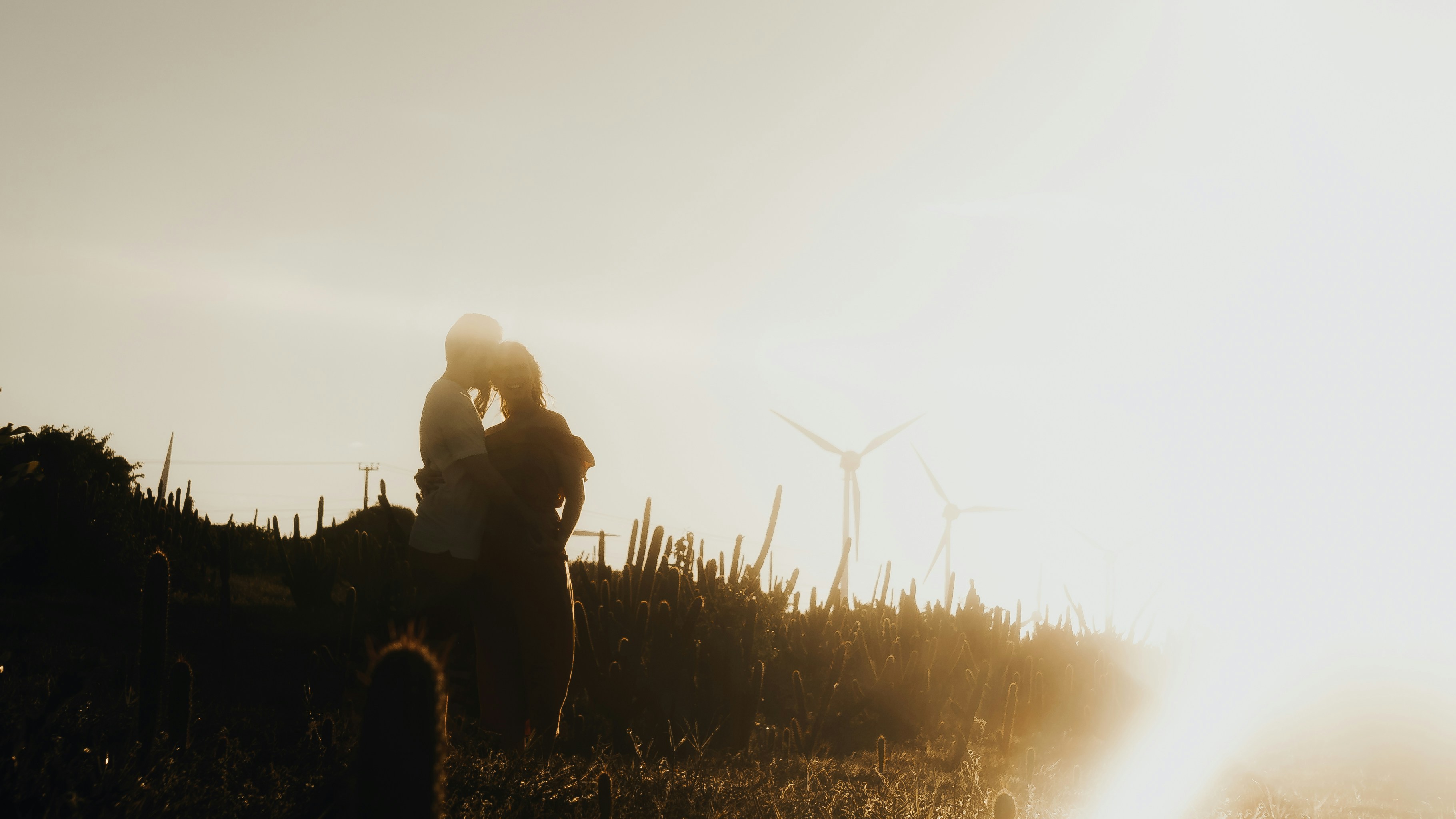 Silhouetted couple sharing a tender moment against a backdrop of wind turbines at sunset.