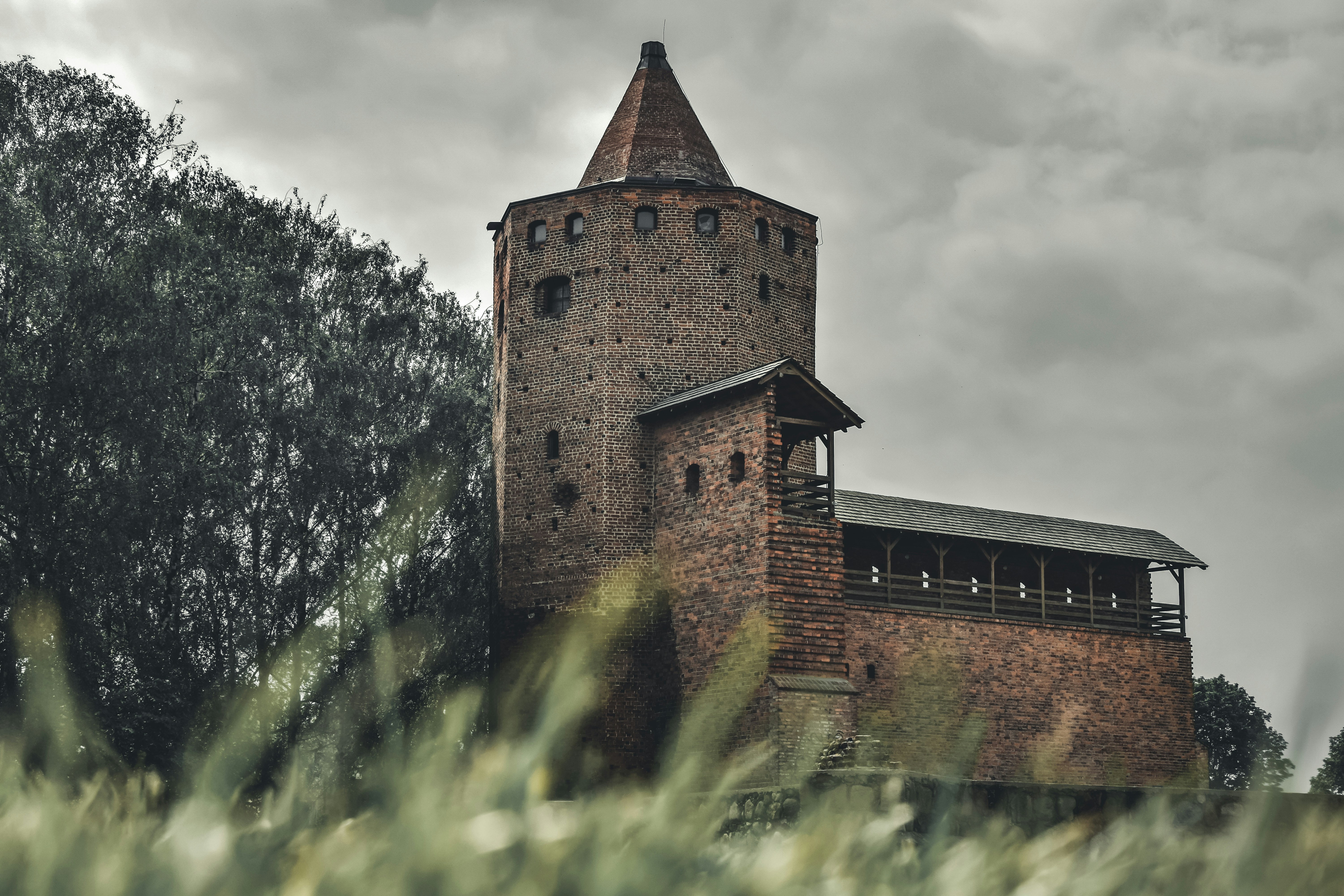Historic brick tower with conical roof set against a cloudy sky, surrounded by lush greenery.