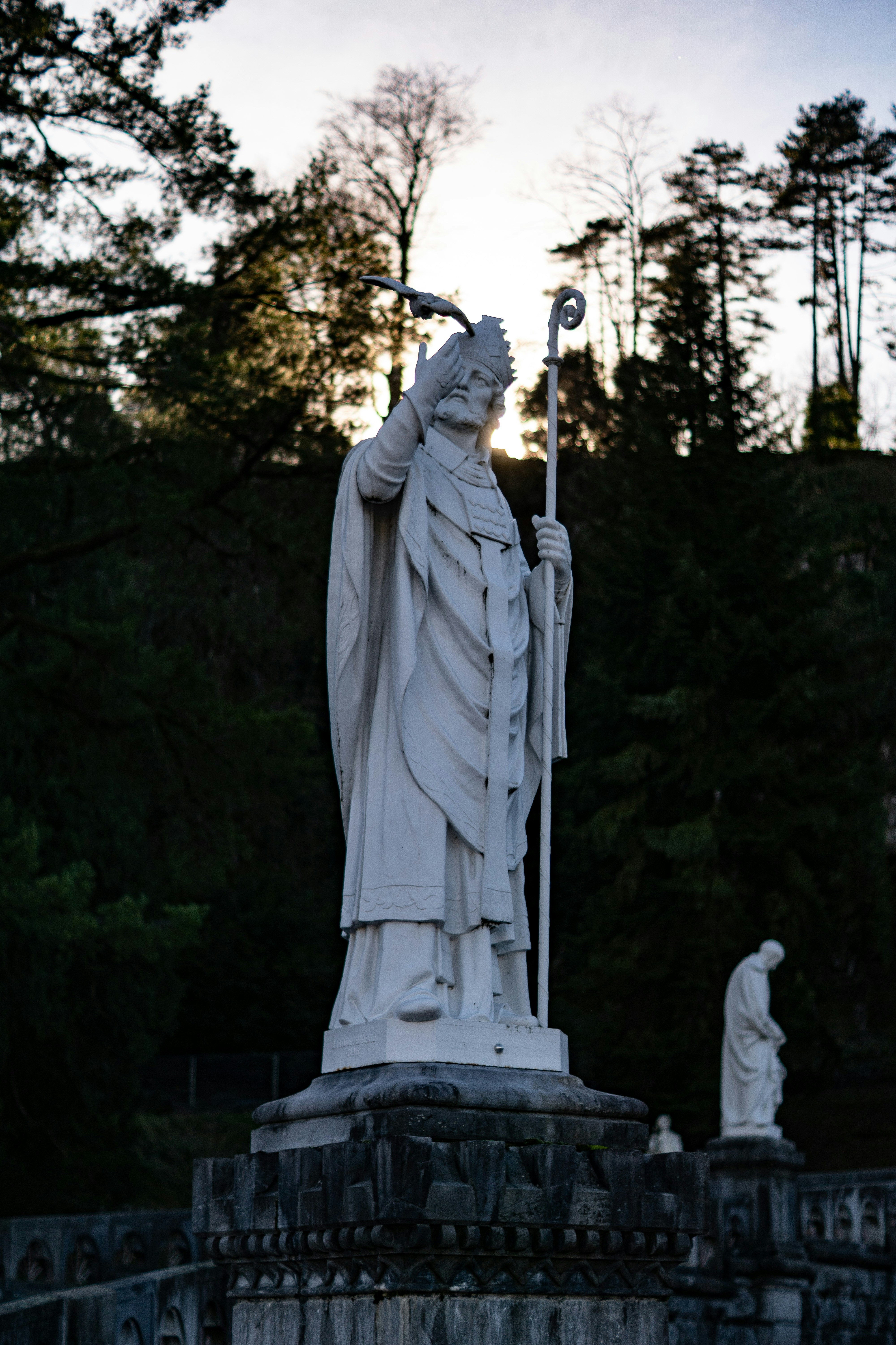 Statue of a robed figure holding a staff, silhouetted against a backdrop of dense trees and a bright sky.