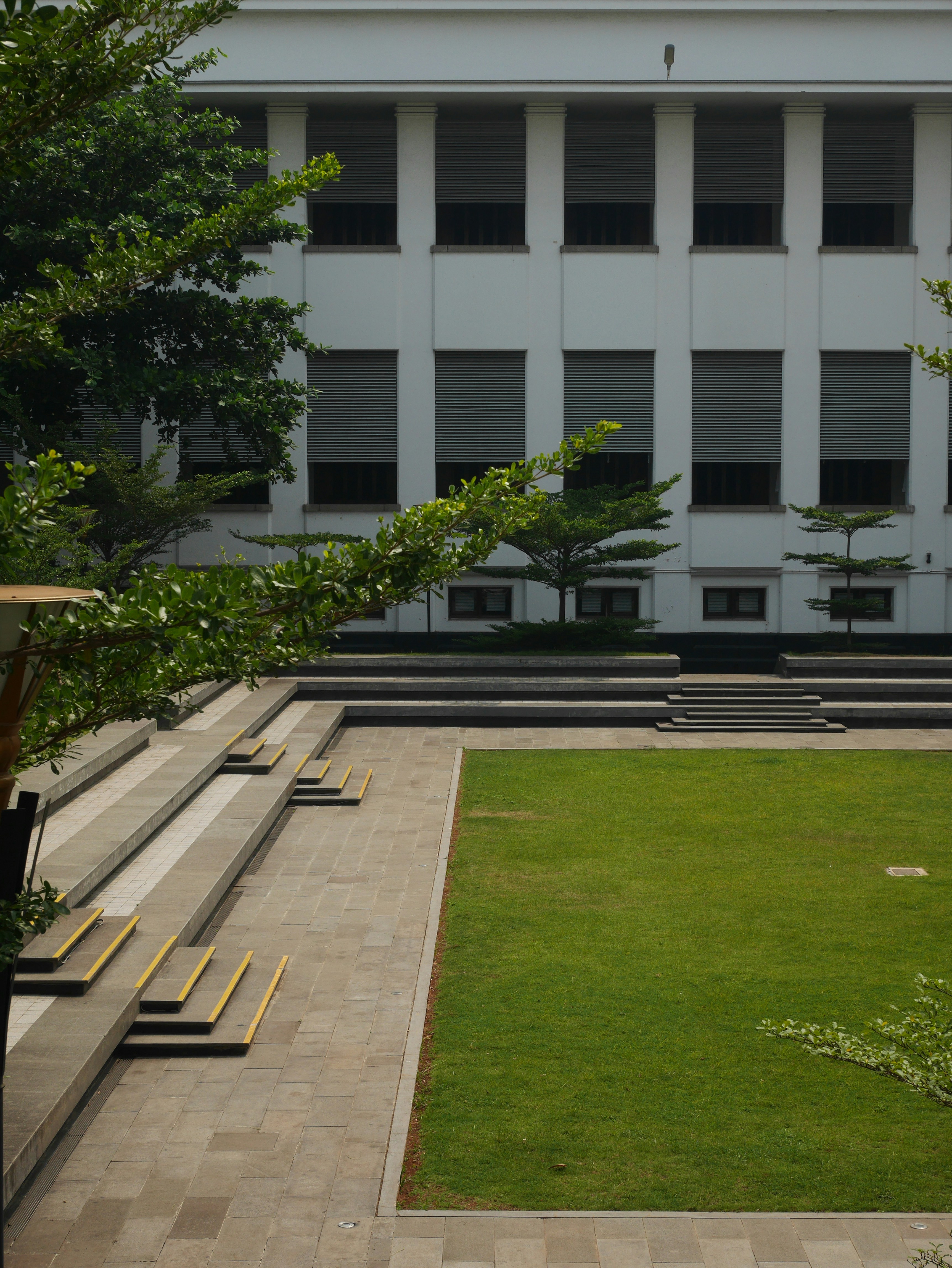 A modernist courtyard with stepped concrete terraces, a trimmed green lawn, and a white façade with tall, shuttered windows.