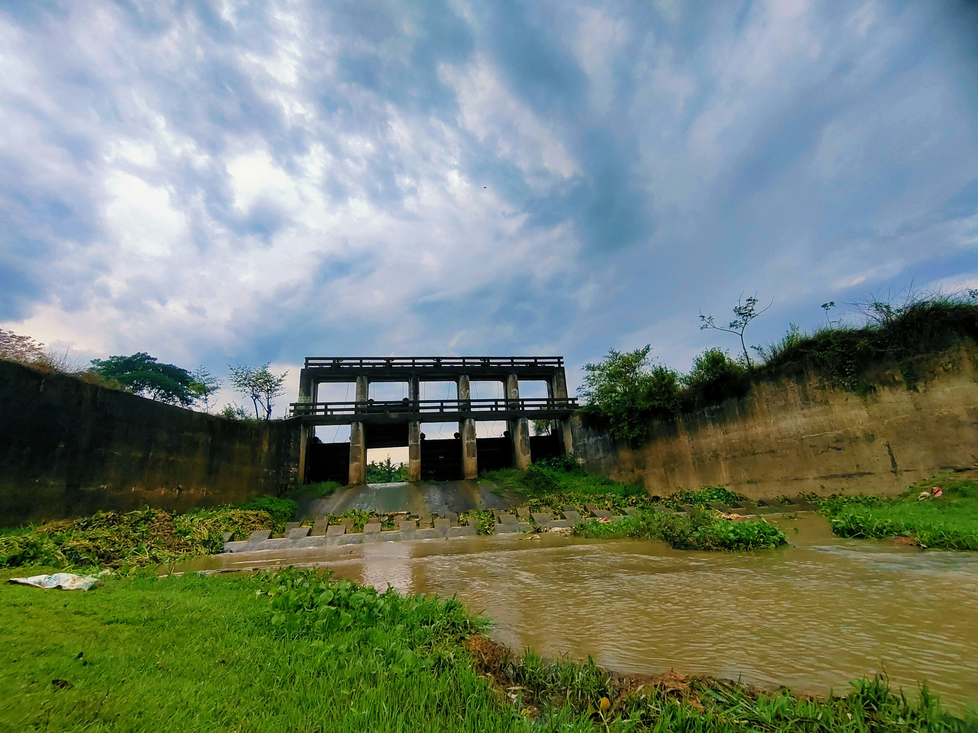 An abandoned gatehouse spans a murky canal, framed by grassy banks and weathered walls under a dramatic, cloud-filled sky.