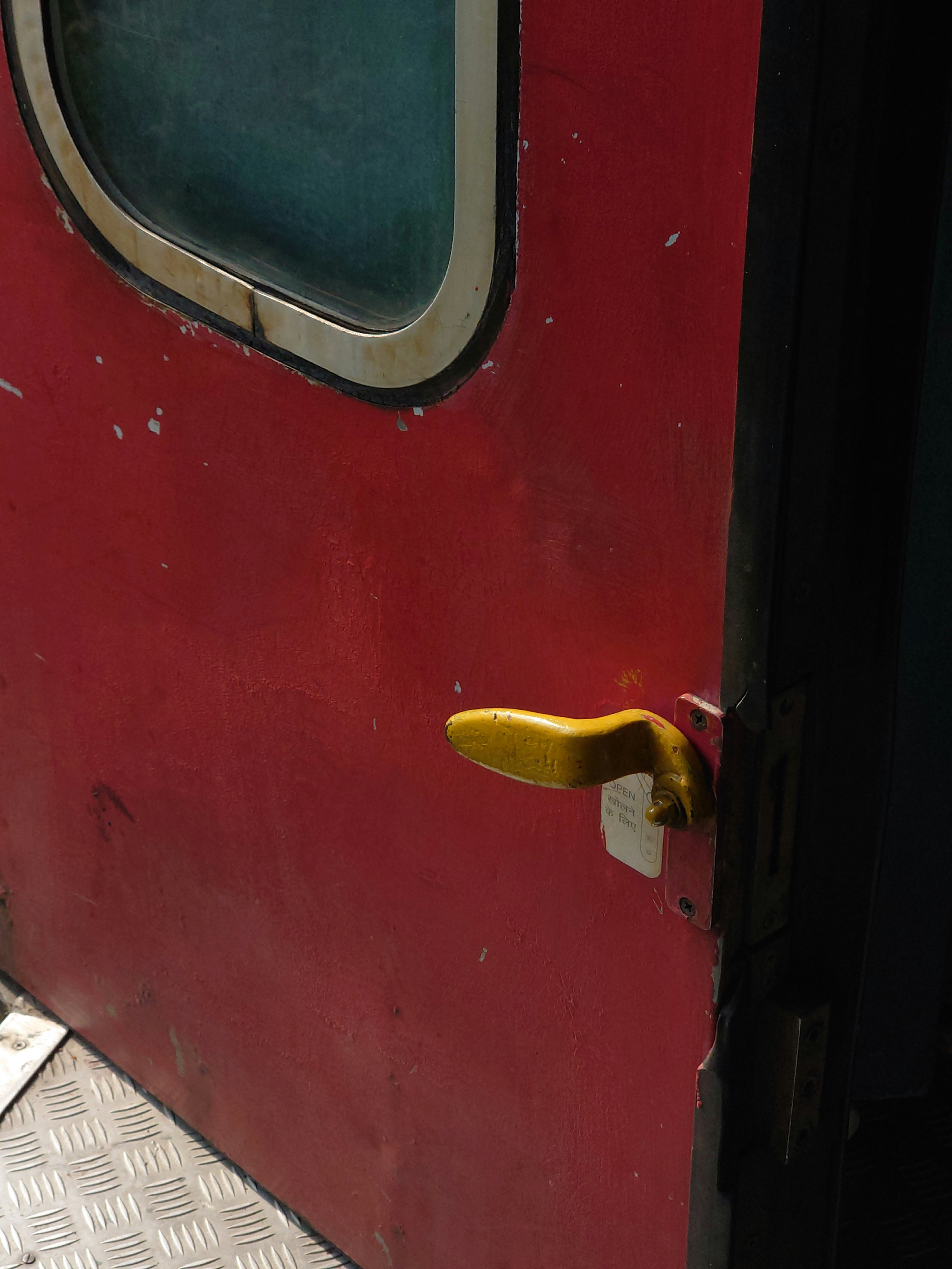 Close-up photograph of a yellow door handle on a weathered red metal door, with a window frame and textured metal floor.