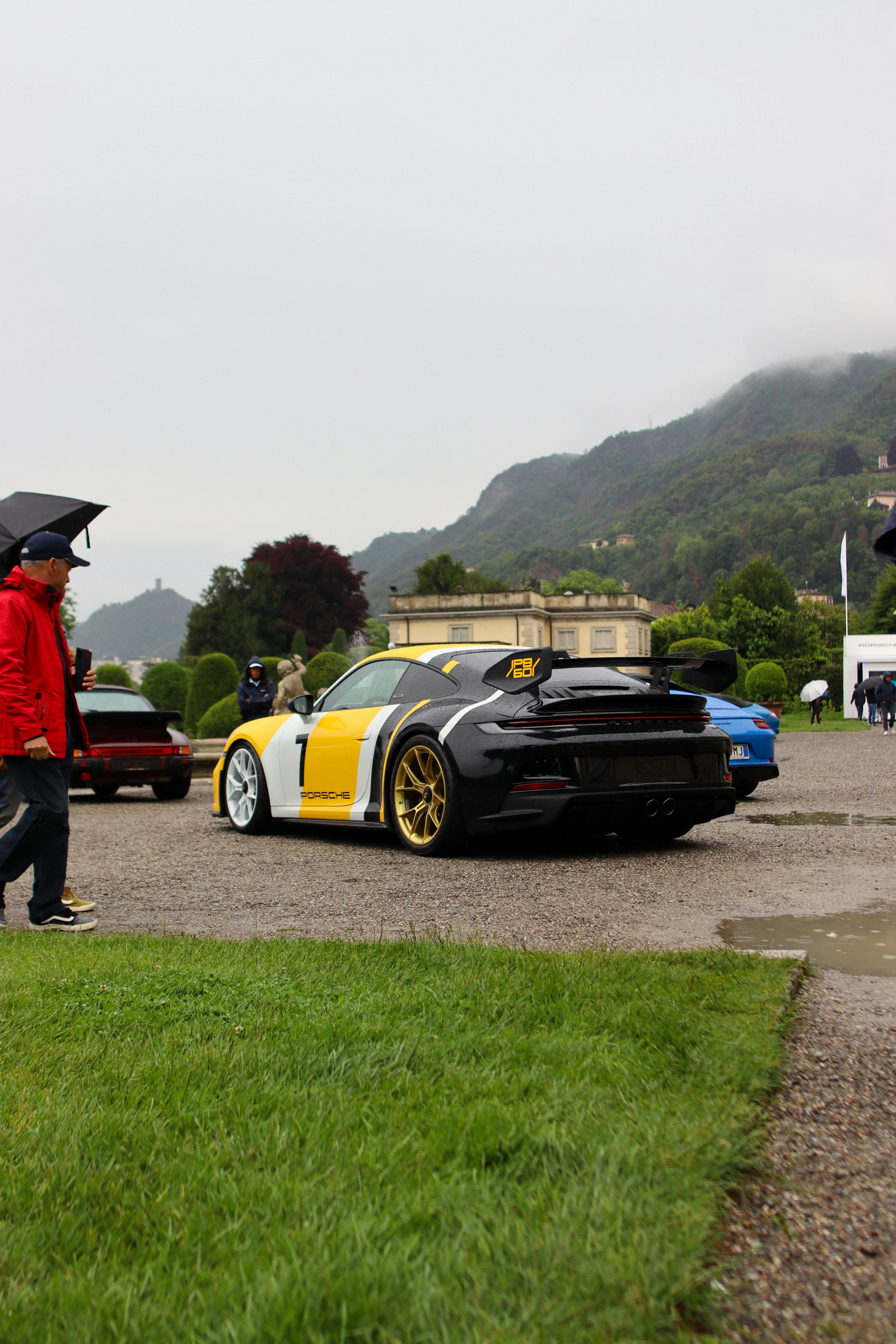 Black Porsche 911 with Yellow Racing Stripe Graces a Gravel Car Show Under Misty Hills