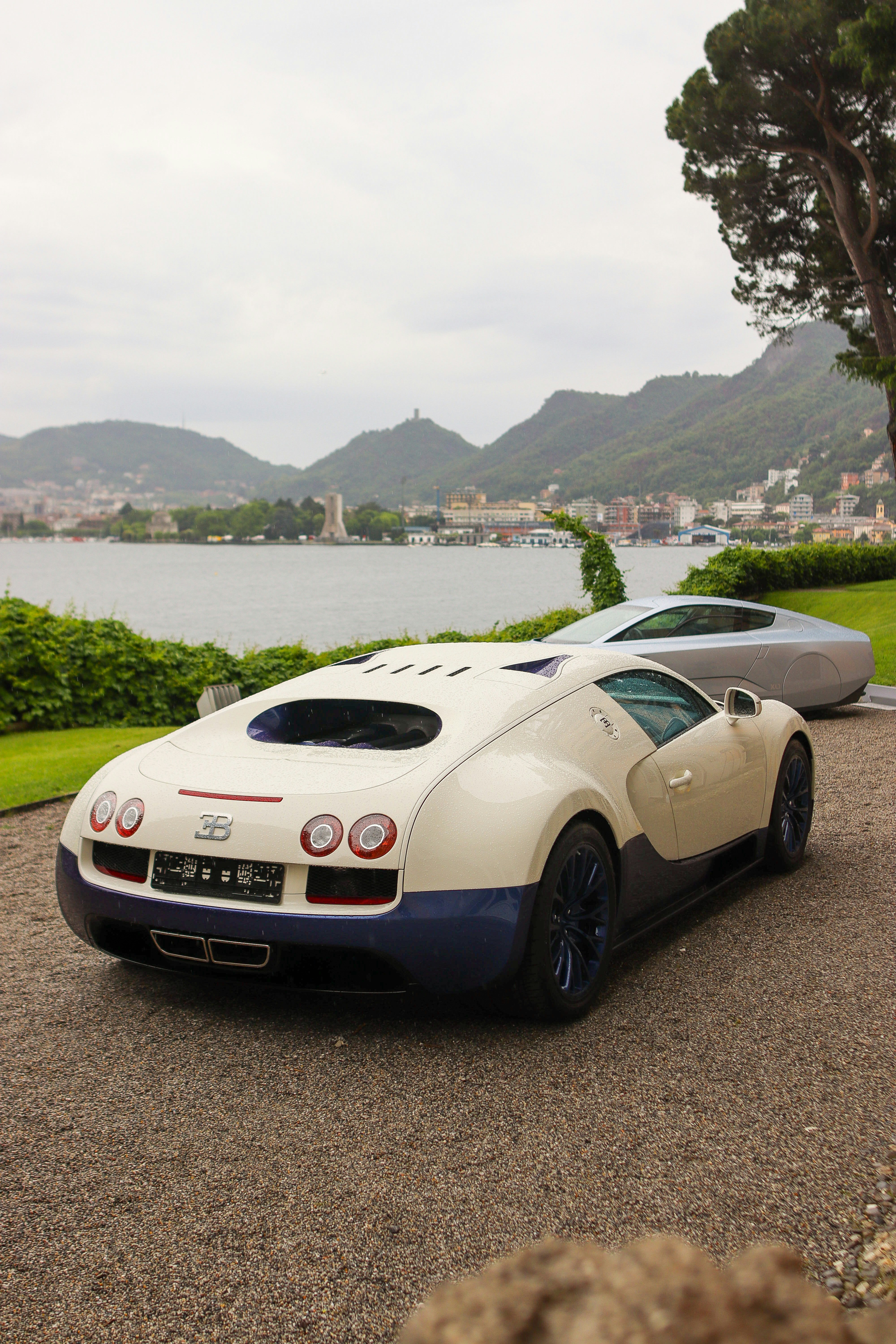 Two luxury sports cars parked on a gravel path with a scenic lake view and mountains in the background.