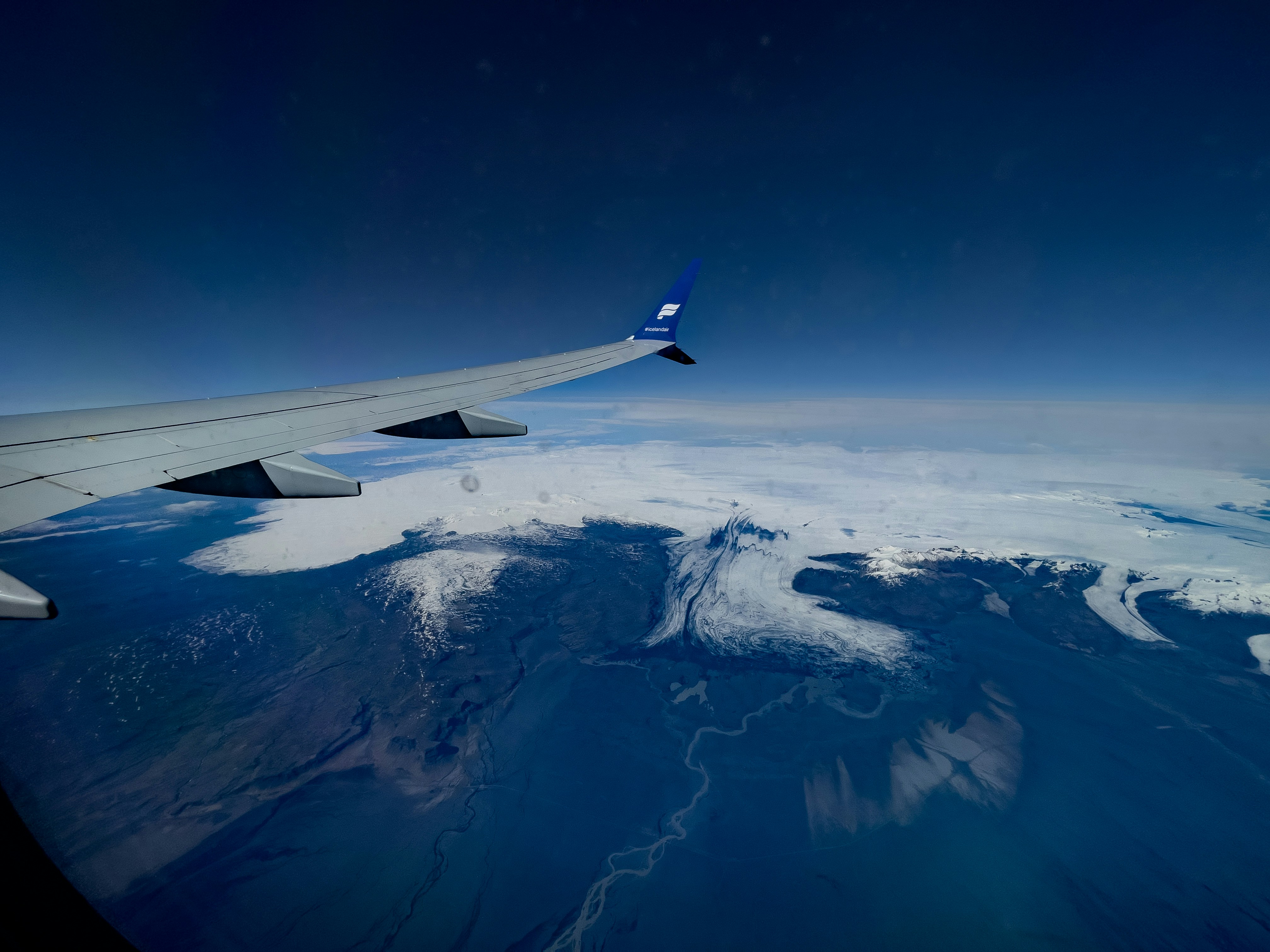 Airplane wing and window view over a cloud-filled ocean at altitude. The photo emphasizes the horizon and atmospheric gradients.