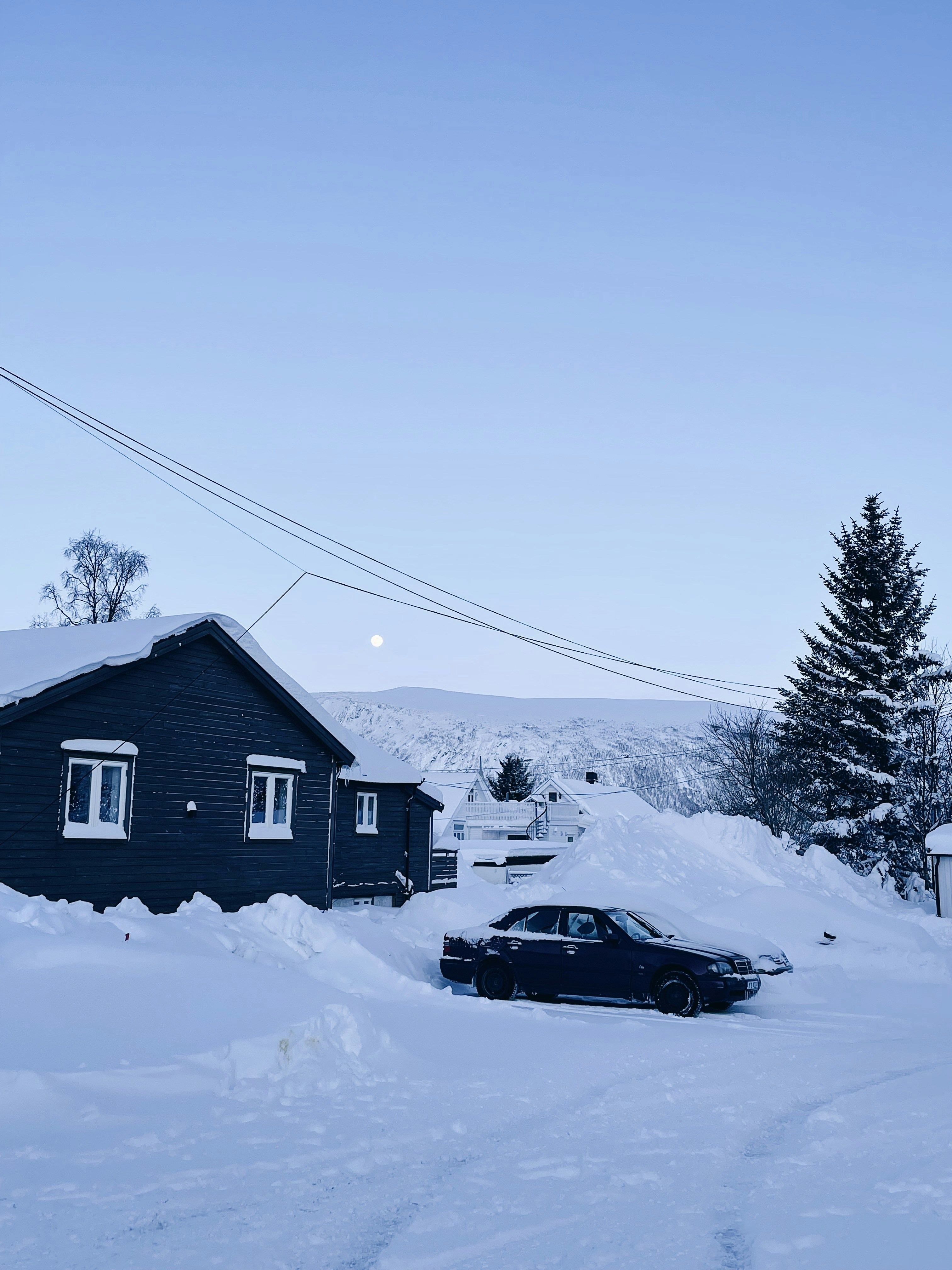 Snow-covered residential scene at blue hour features a dark wooden house, a parked black car, and piled drifts beneath a clear sky with the moon visible.