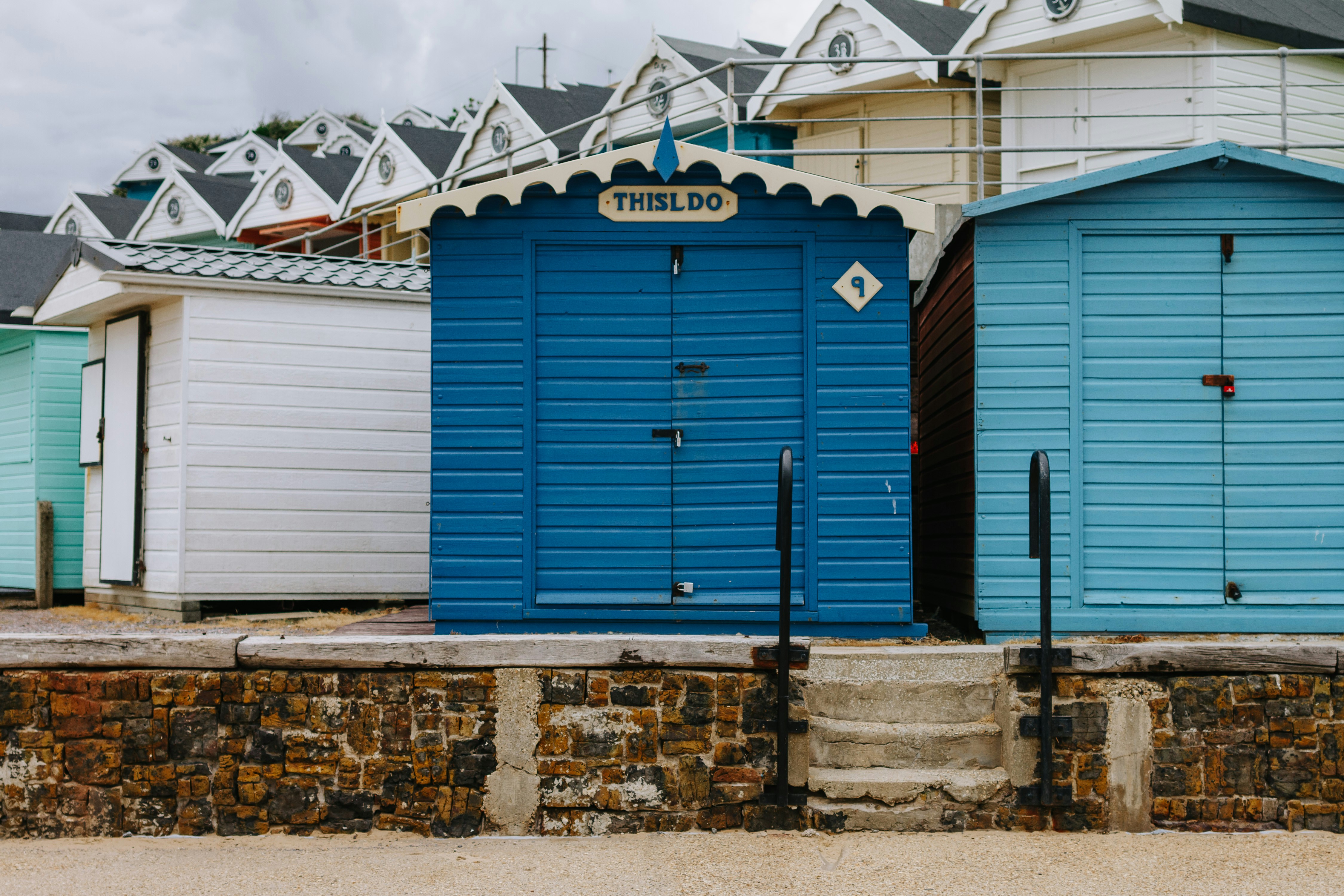 Blue beach hut nestled between pastel-colored neighbors on a cloudy day.