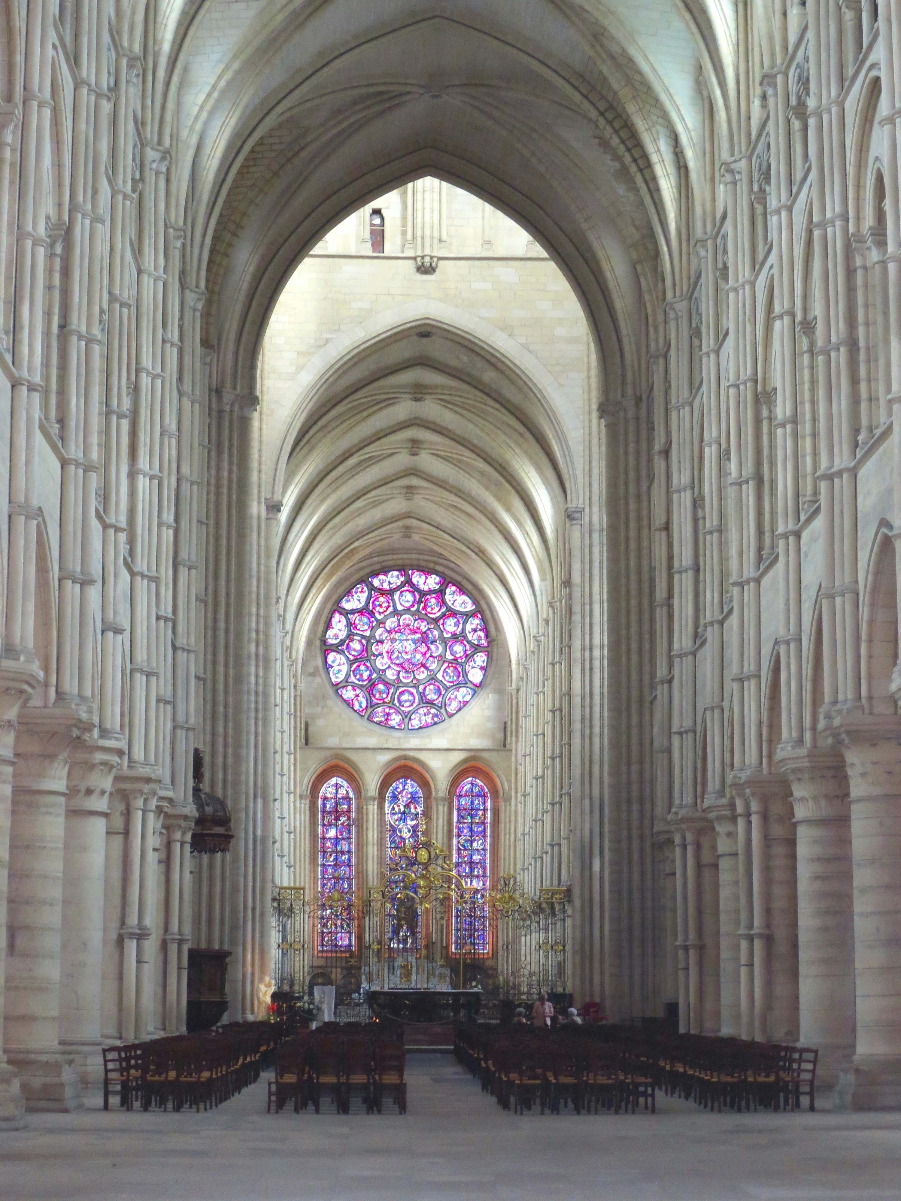 Interior of a Gothic cathedral with soaring arches leading to a vibrant rose window and stained-glass panels behind the altar.
