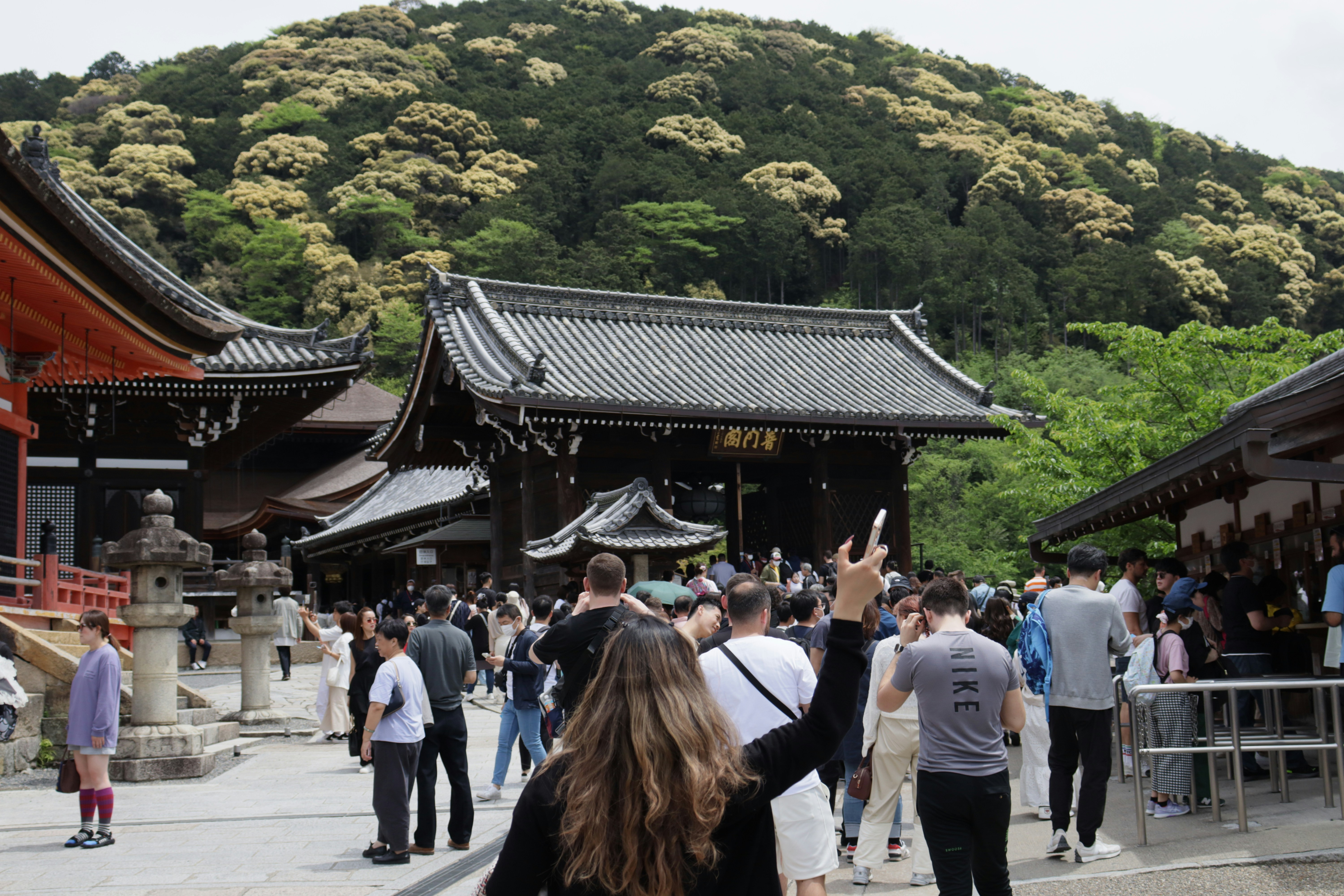 Crowd gathers at a traditional temple surrounded by lush greenery and a hill dotted with blooming trees.