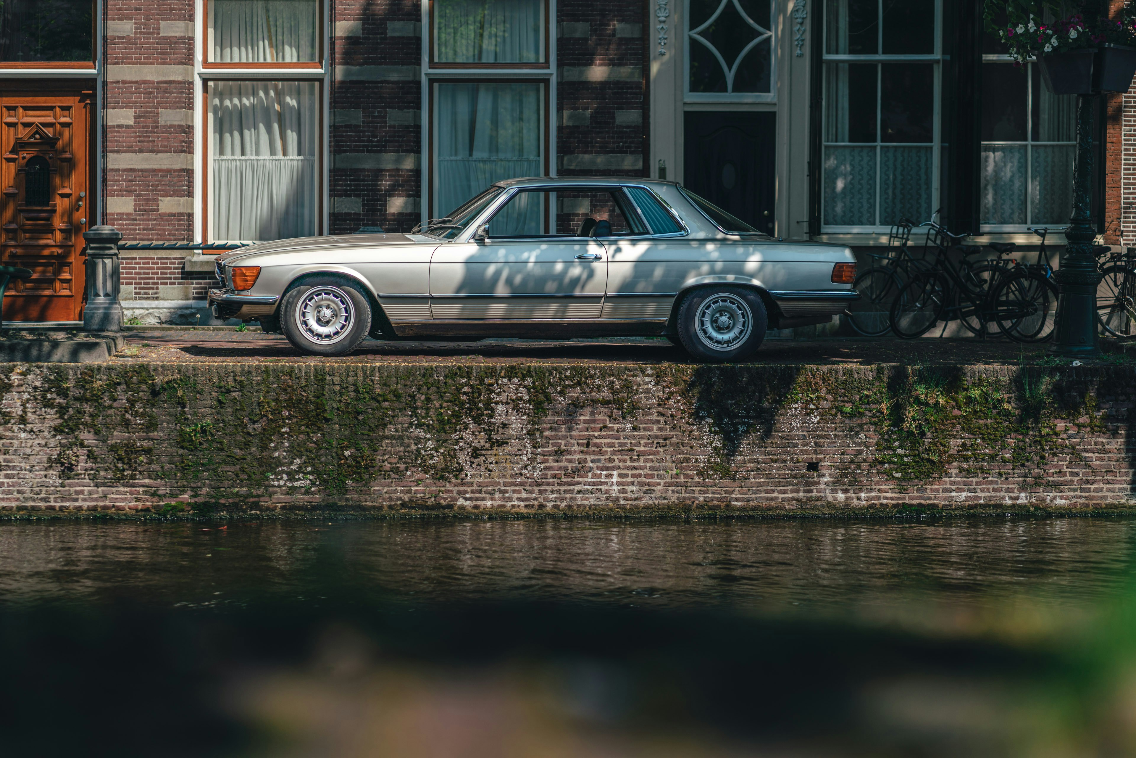 Silver classic car parked beside a tranquil canal, framed by a brick building facade and dappled sunlight.