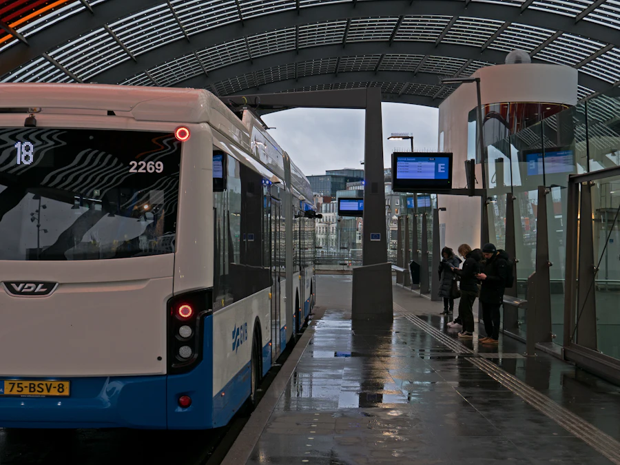 A city bus is parked under a curved glass-and-steel transit canopy on a wet platform. A small group of people waits near the right side beside glowing departure signs.