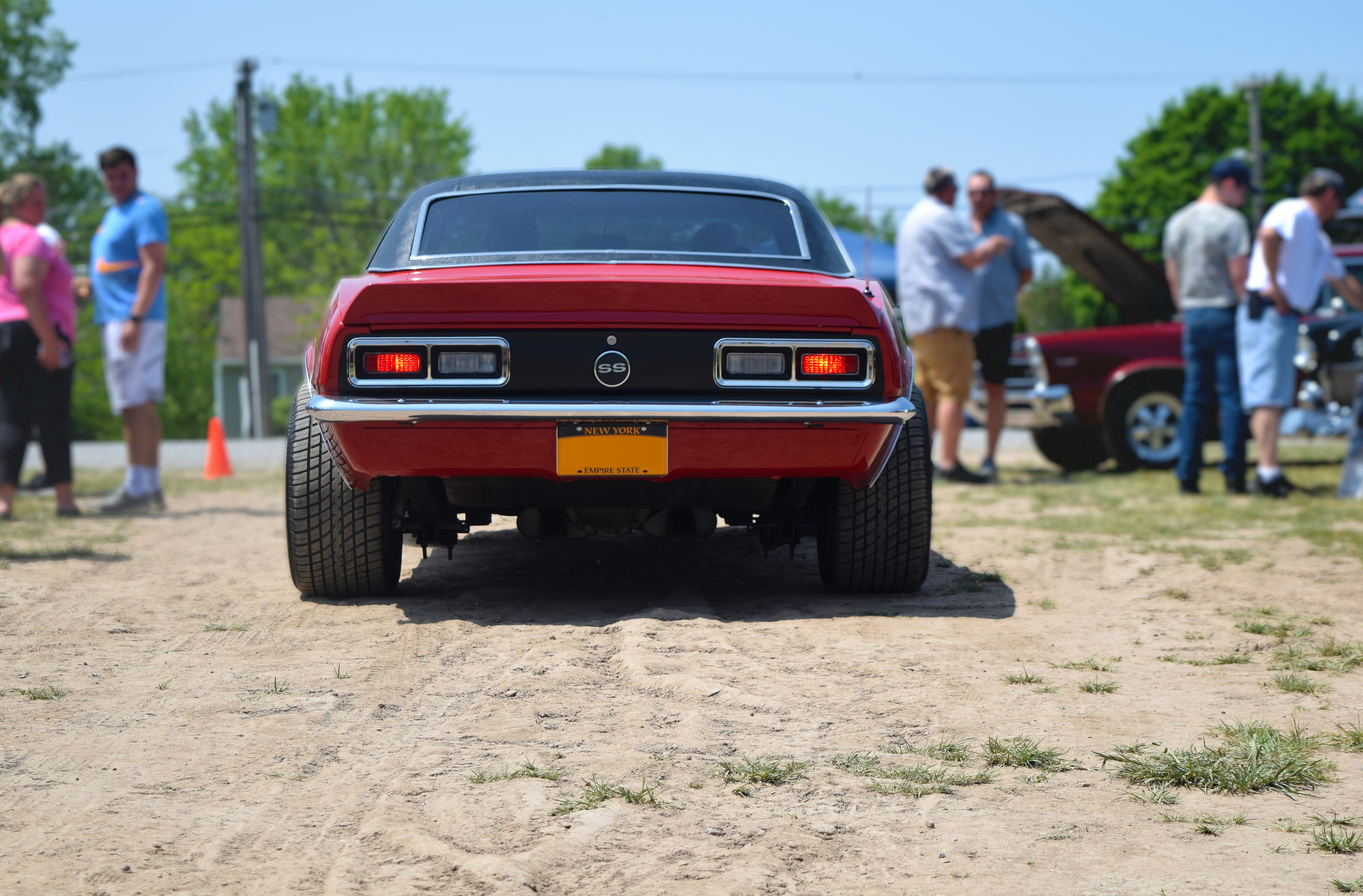 Rear view of a vintage red car parked on a dirt road with people gathered nearby on a sunny day.