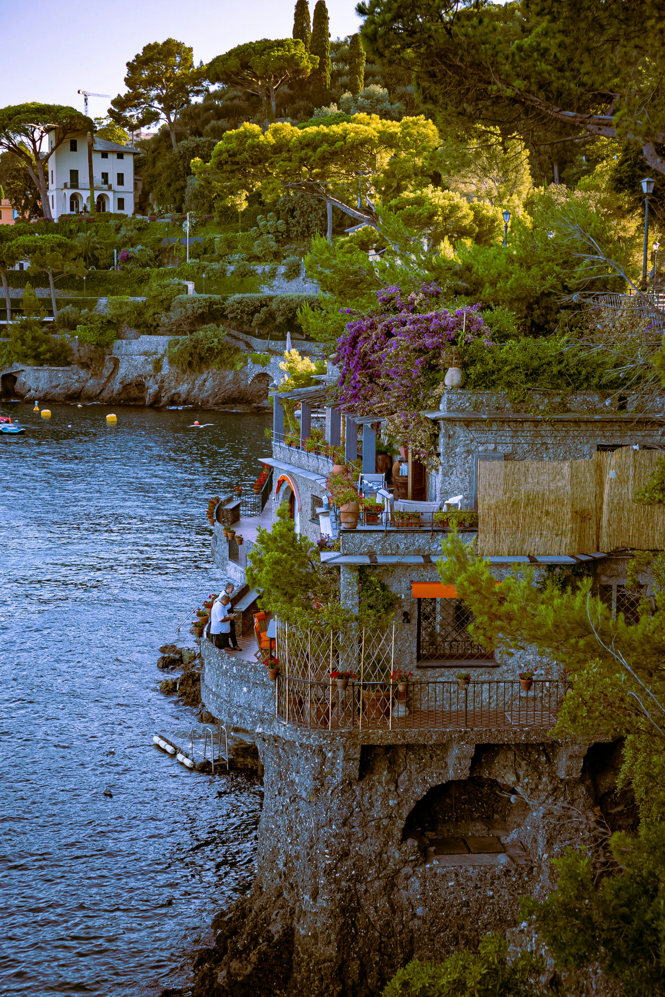 Charming coastal structure adorned with vibrant bougainvillea, overlooking a tranquil bay with kayakers. The scene captures the essence of leisurely seaside living.