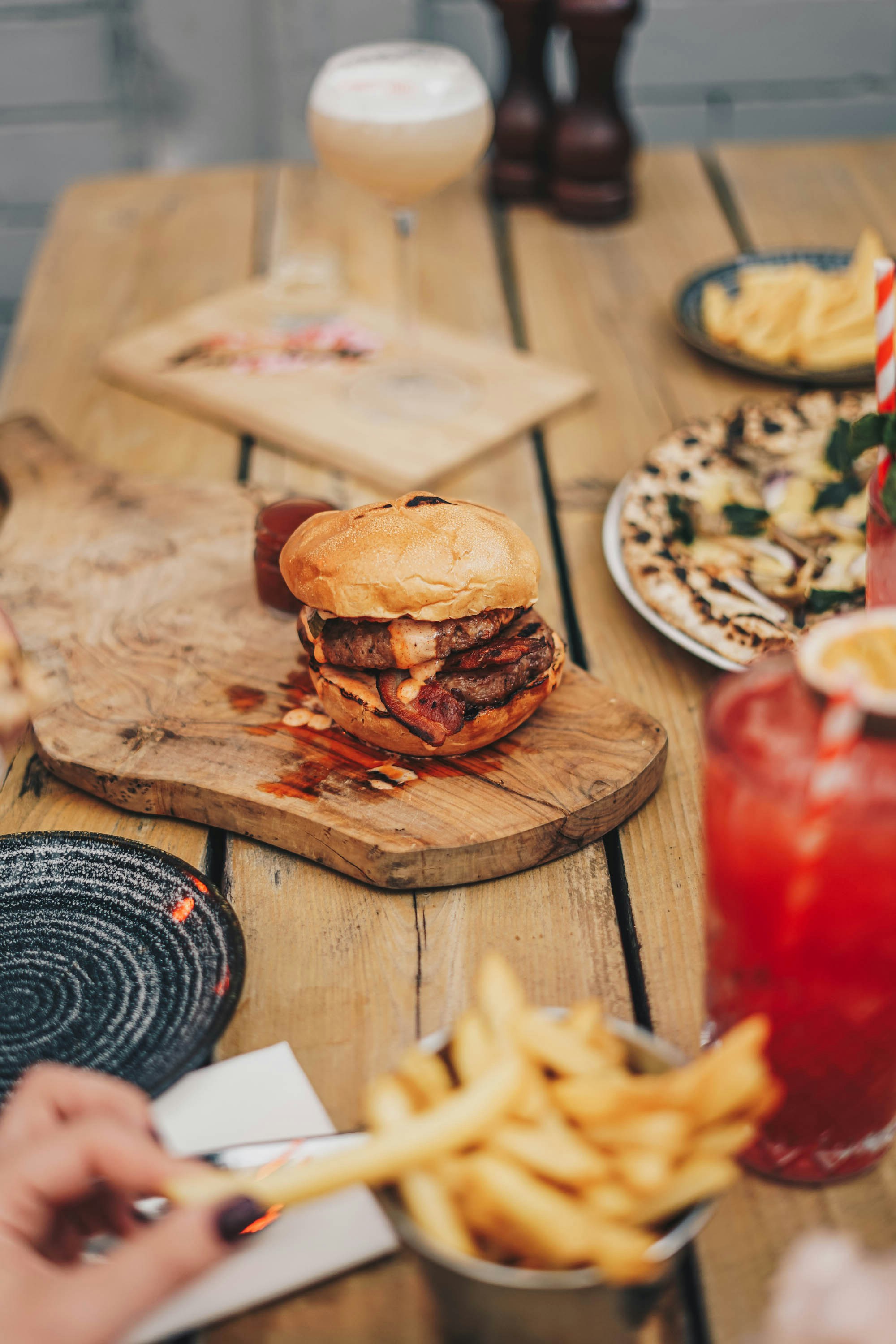 Sunlit Burger on a Wooden Board: Fries, a Crimson Drink, and a Pizza Plate in the Background