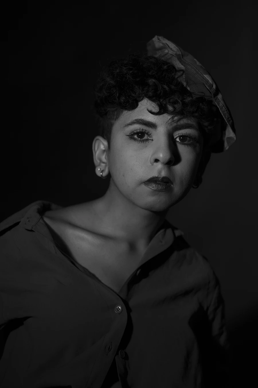 Black-and-white portrait of a person with curly hair and a tilted hat, lit dramatically against a dark background. The subject faces the camera with a calm, serious expression.