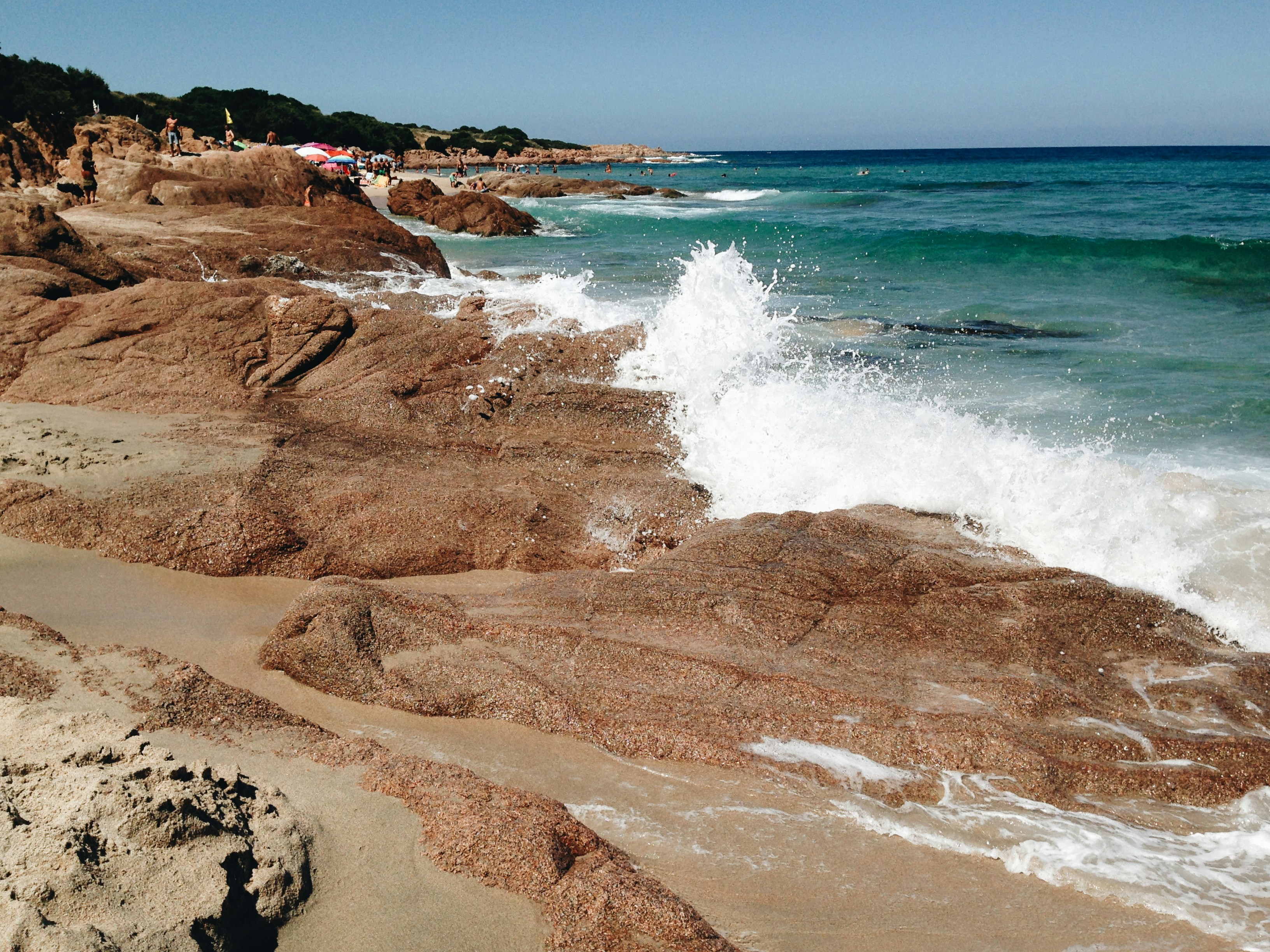 Sunlit rocky coastline with turquoise water and white-capped waves rolling onto the shore; a small beach scene with distant people under clear skies.
