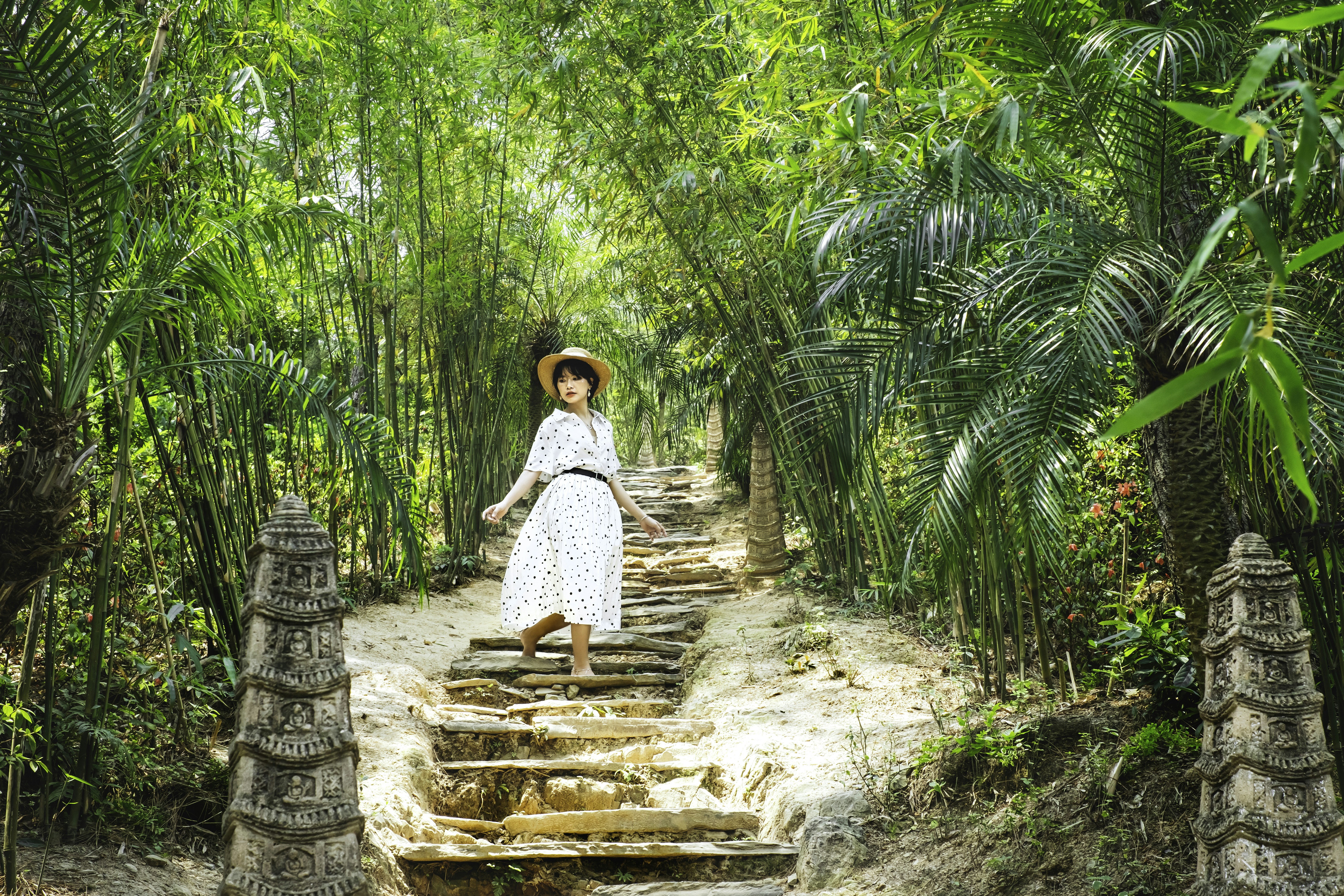 A woman in a polka dot dress strolls down a stone path surrounded by vibrant greenery and ancient stone sculptures.