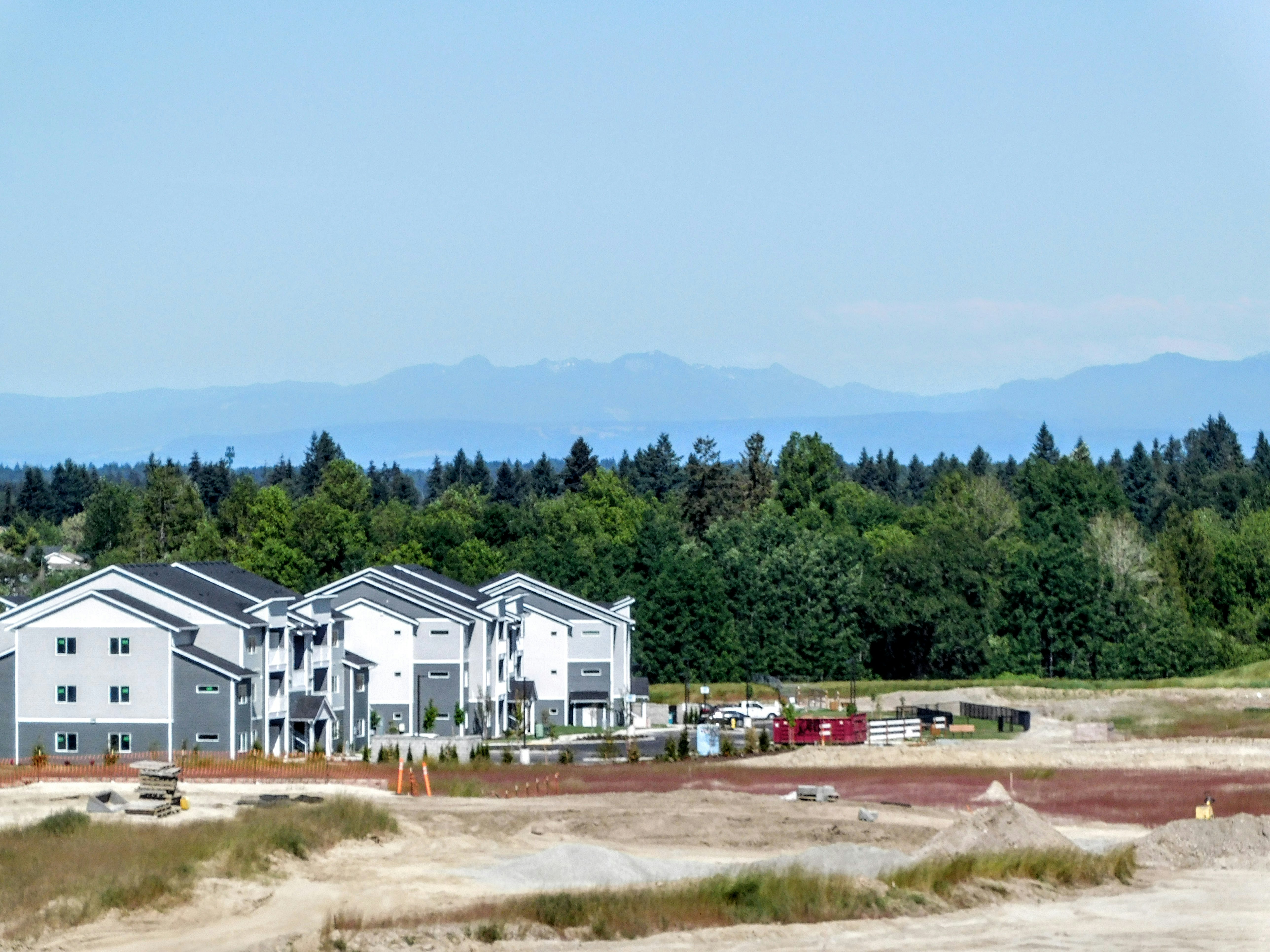 Photograph of a row of modern white townhomes under a clear blue sky, with red dirt foreground from ongoing construction and a distant tree line.