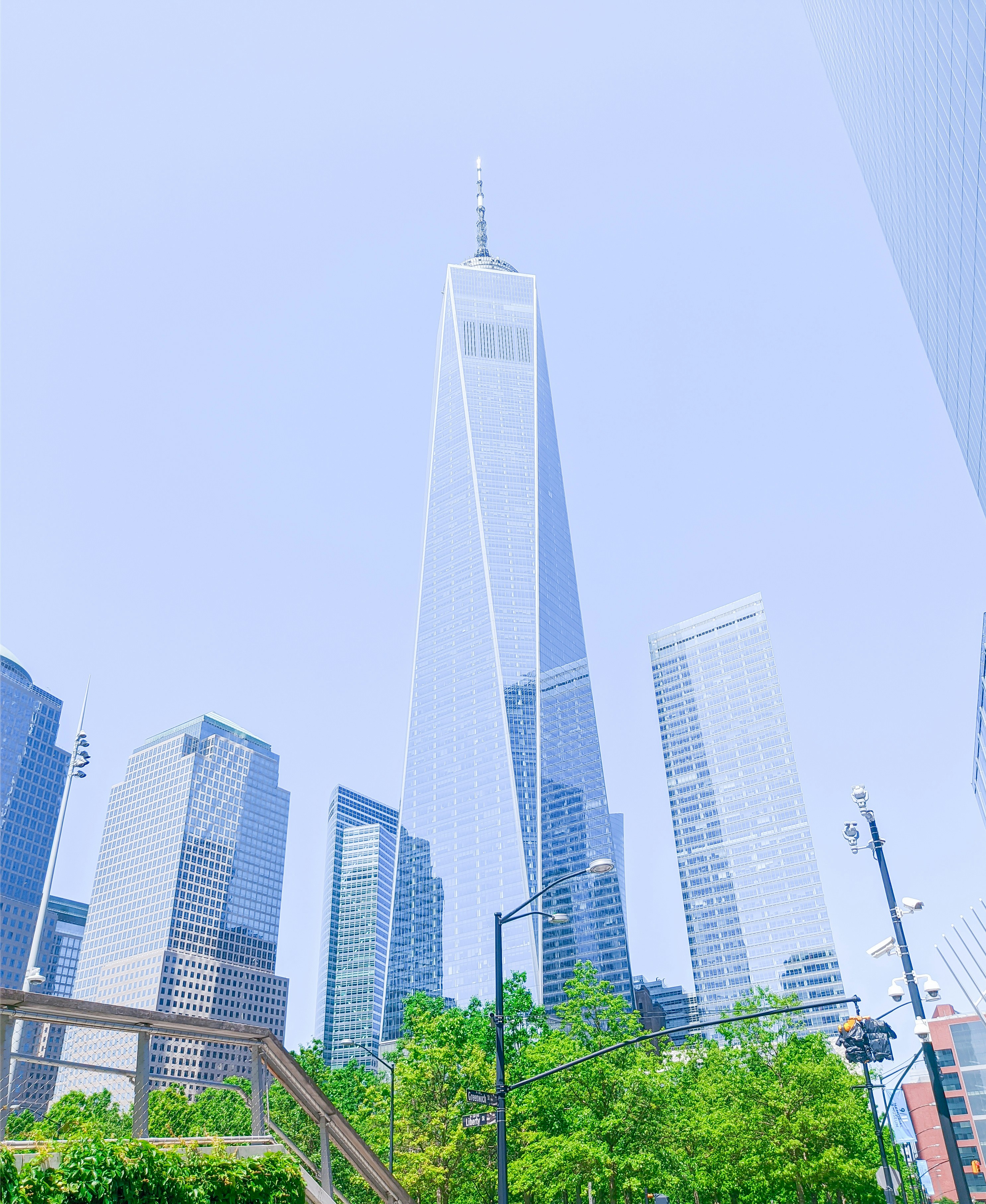 One World Trade Center towers over a blue cityscape with green trees in the foreground under bright daylight.
