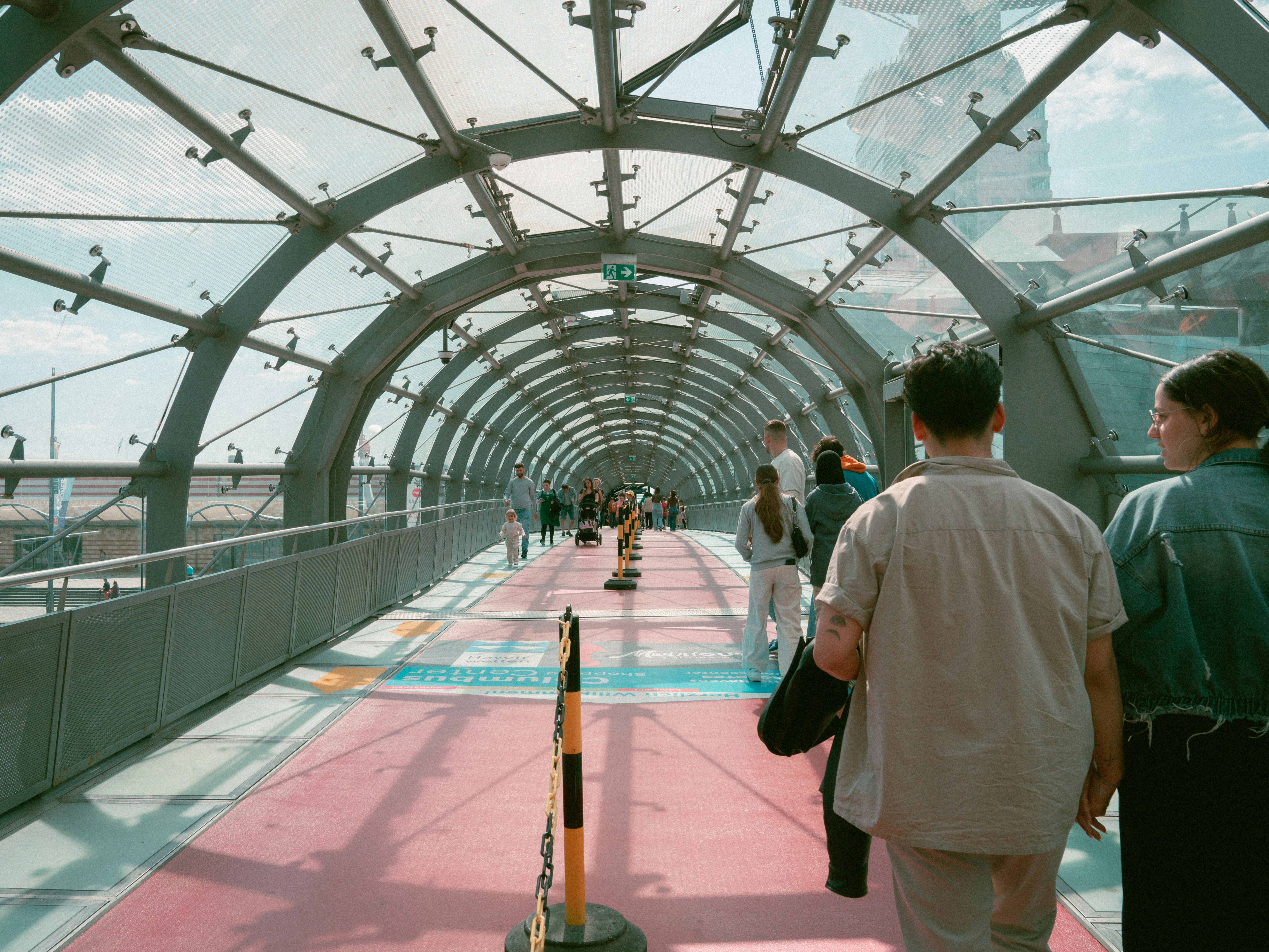 People walking through a glass-encased tunnel connecting two buildings under a clear sky.