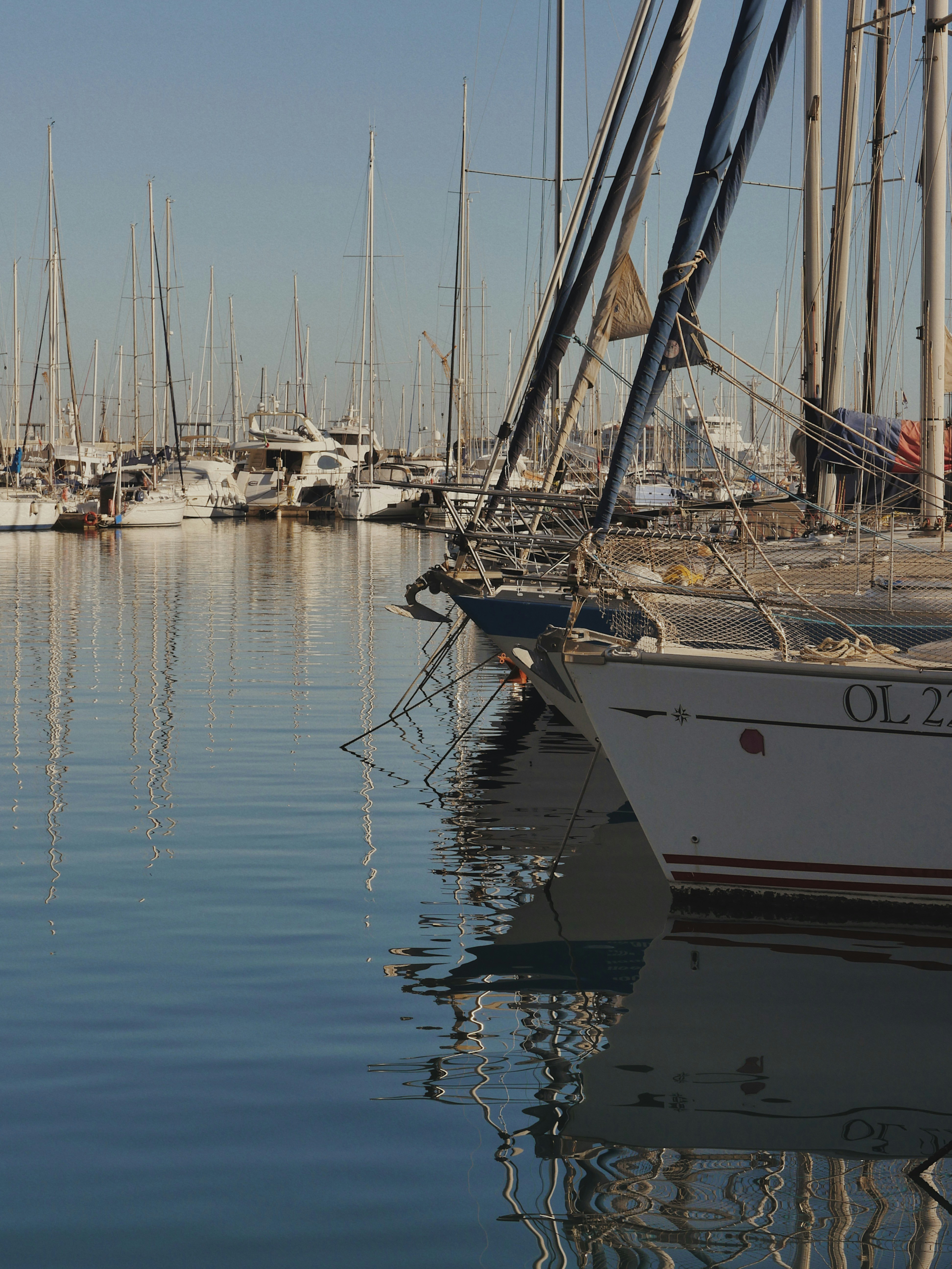 Sailboats moored in a calm marina, their reflections mirrored on the still water under a clear blue sky.