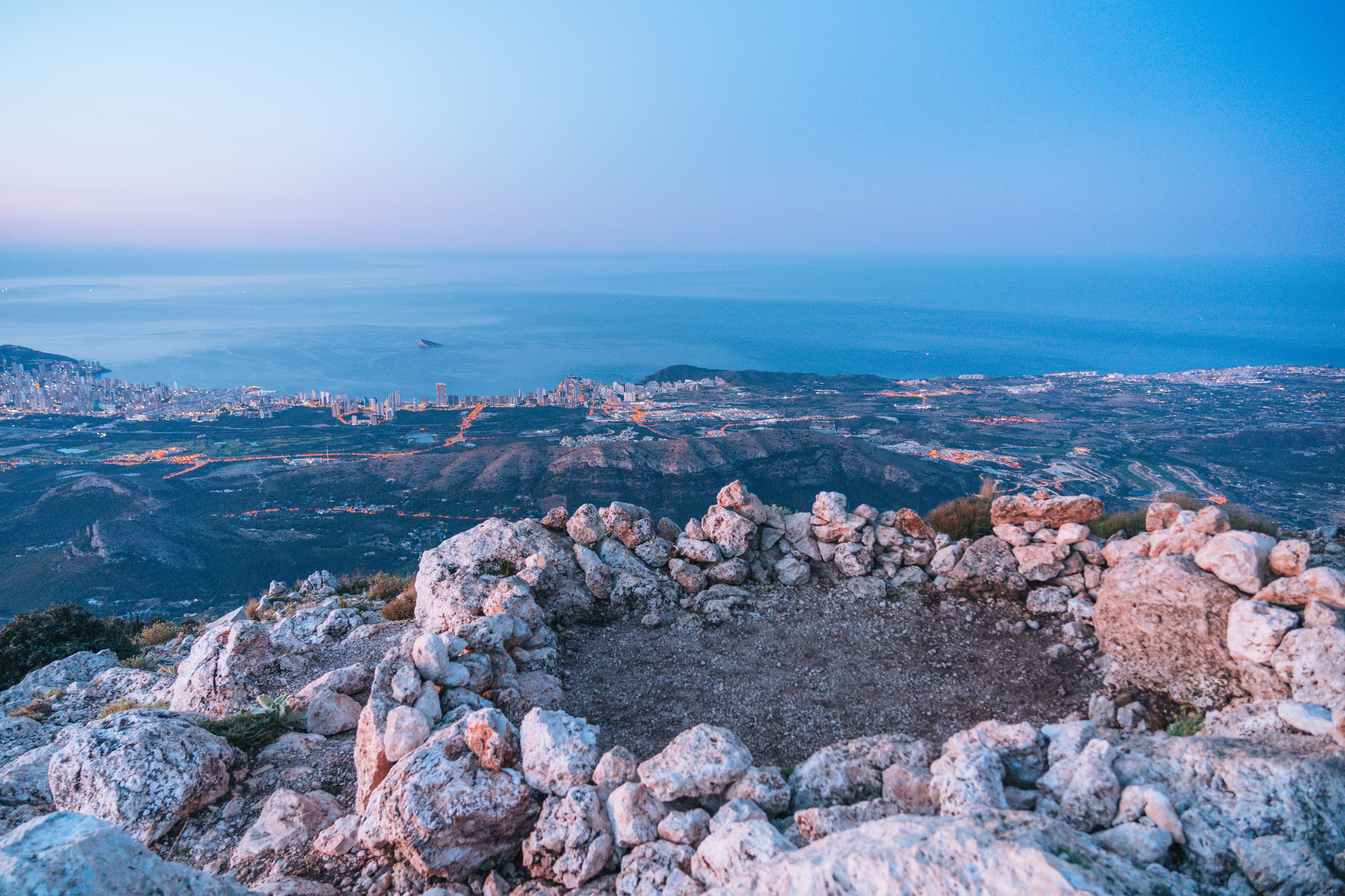 Rocky outcrop overlooking a vast landscape with distant city and sea under a twilight sky.