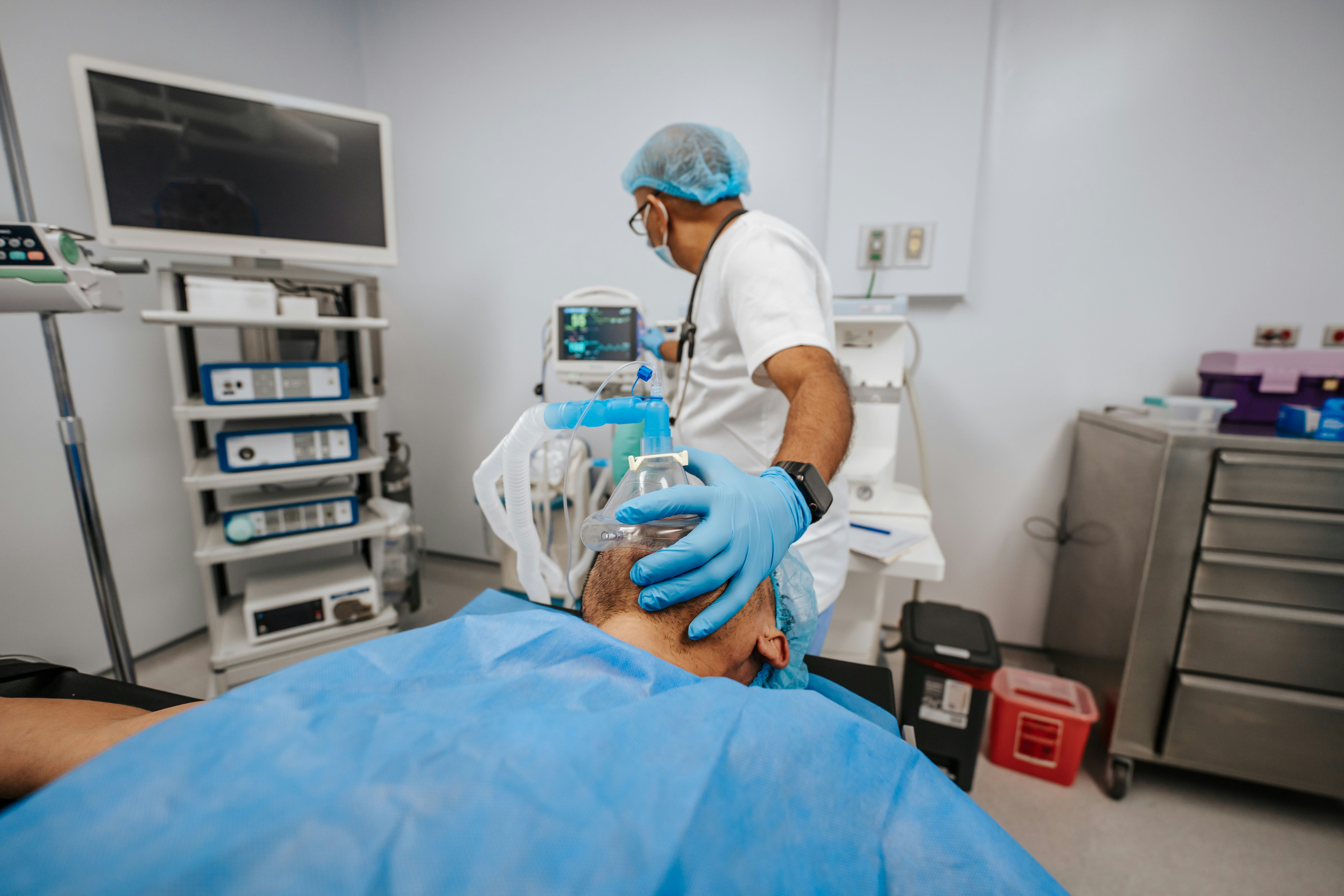 Dentist in a clinical setting checks a patient's teeth, surrounded by medical equipment.