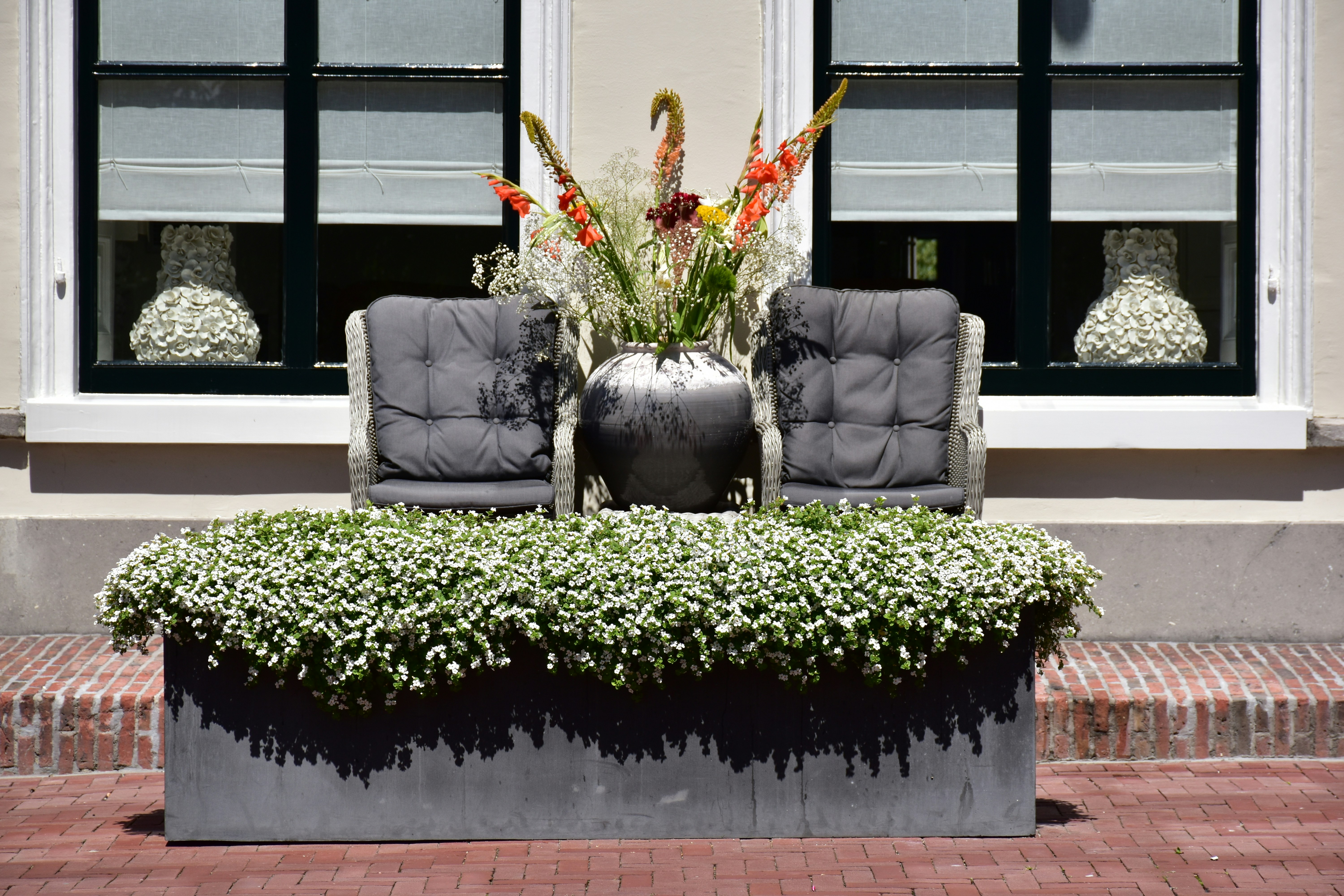 Two cushioned chairs flank a lush floral arrangement in front of a large window with white blooms below.