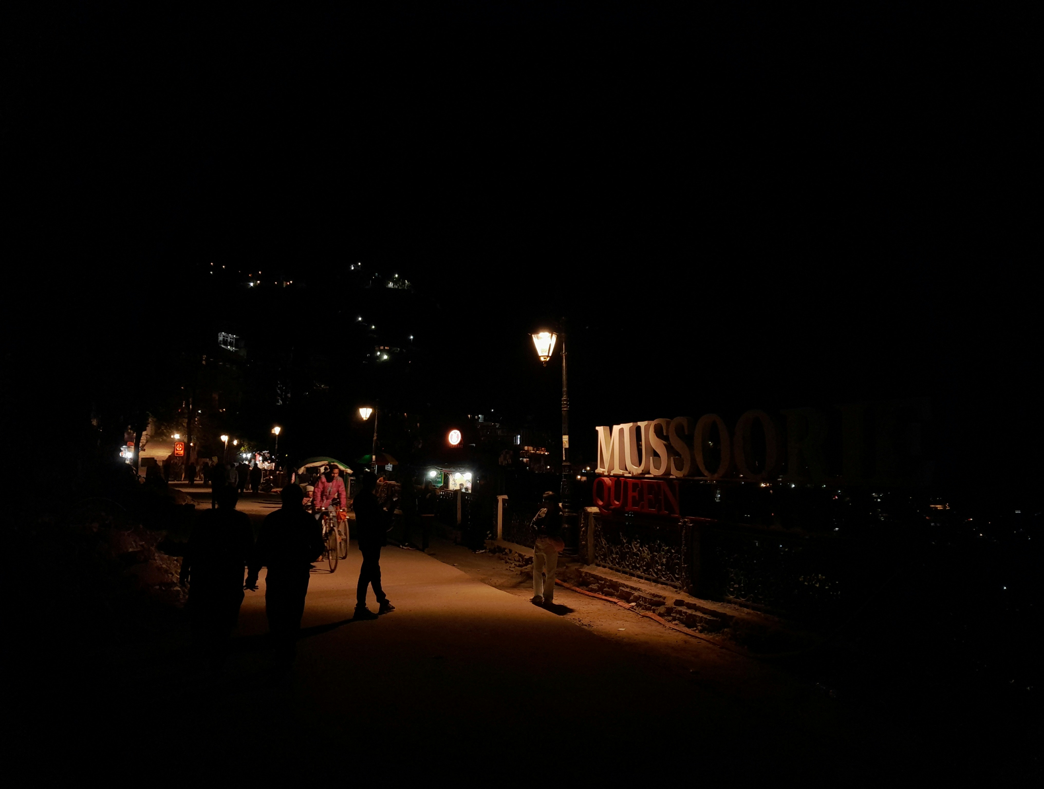 Silhouetted pedestrians walk along a dimly lit promenade under warm street lamps. A distant sign glows softly to the right.
