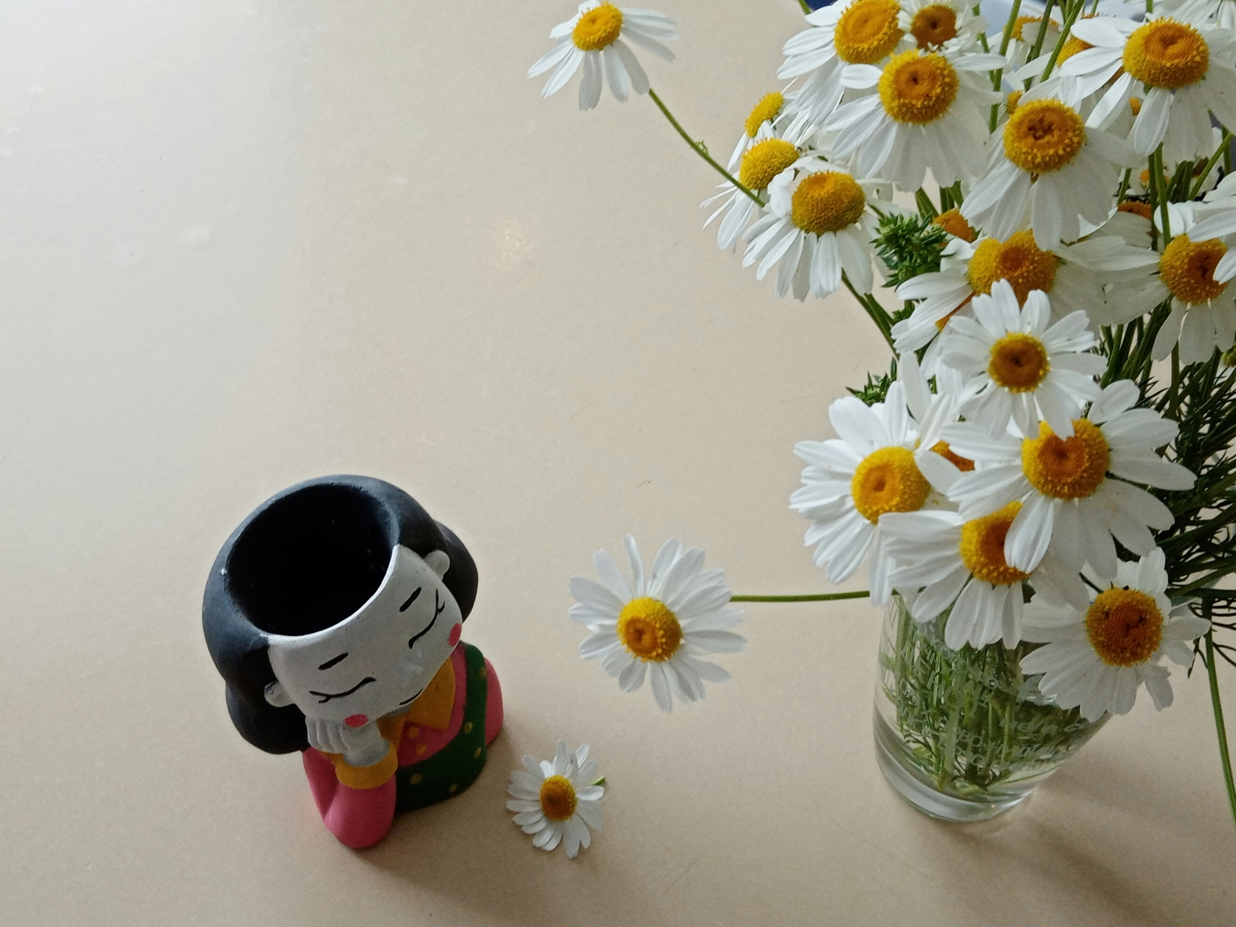 Small ceramic figurine beside a glass vase of daisies in soft light, creating a gentle still life with a playful, everyday mood.