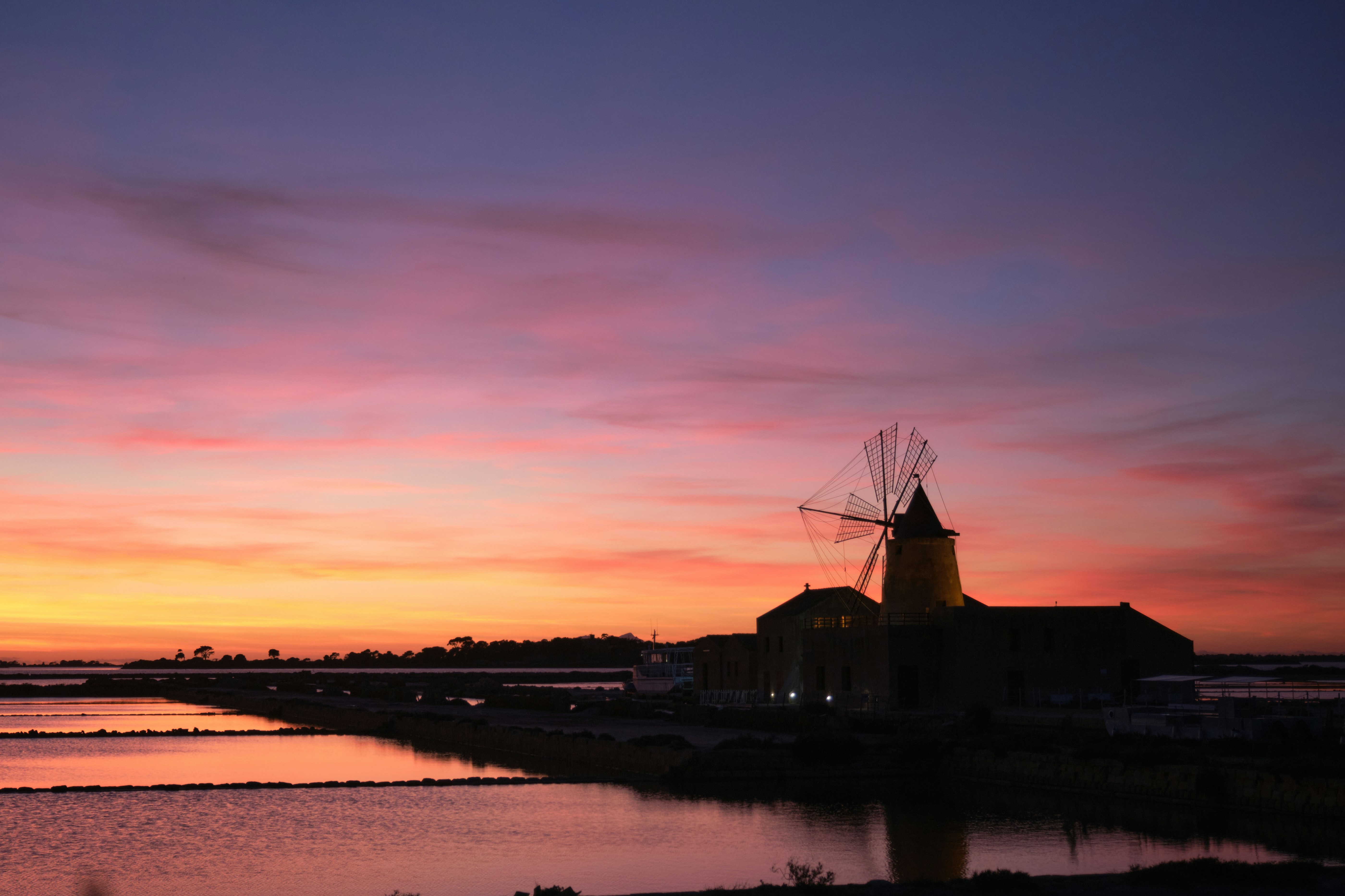 Windmill silhouetted against a vibrant sunset sky reflecting on a calm body of water.