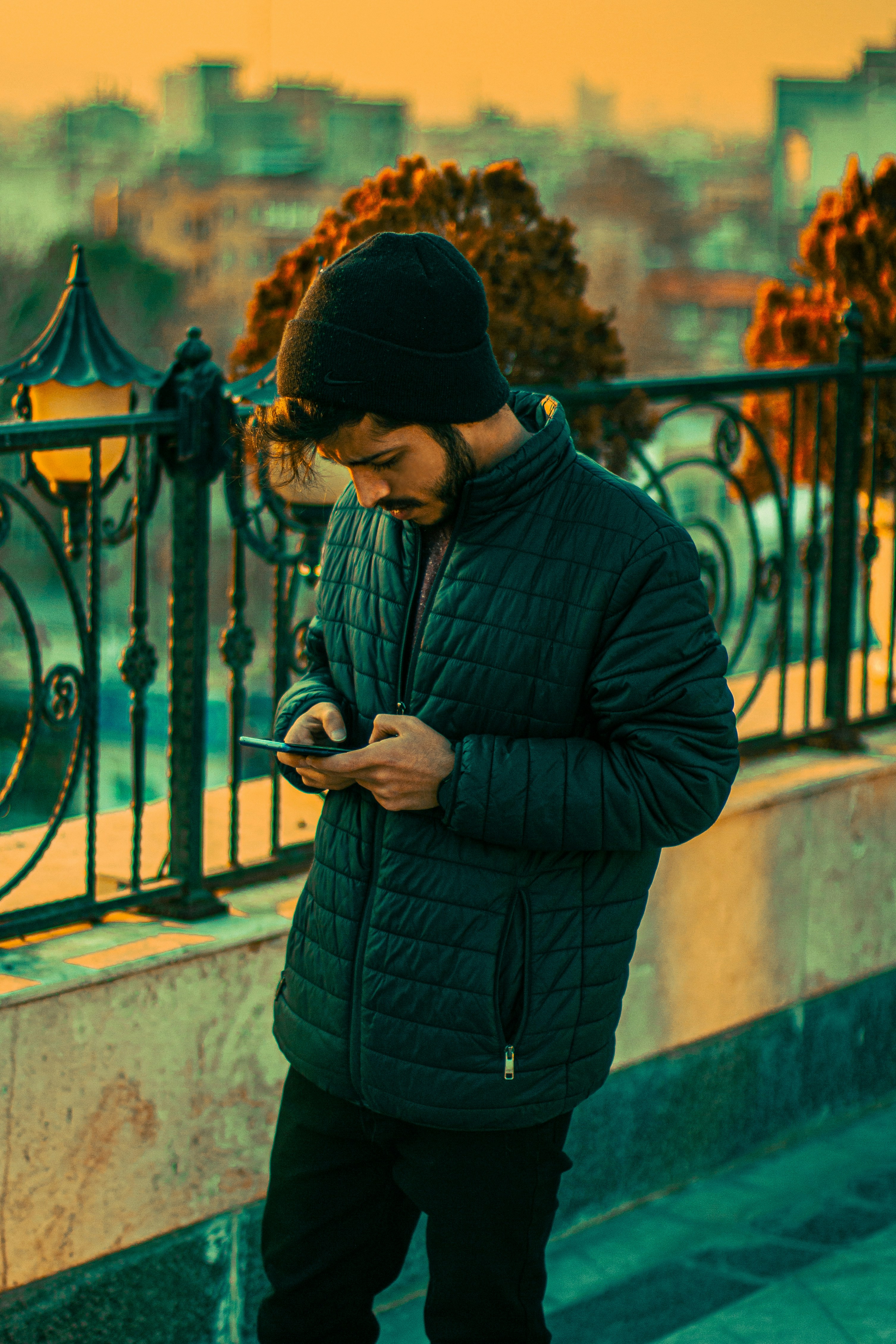 Rooftop Quiet: A man in a teal puffer glances at his phone as evening city lights glow.