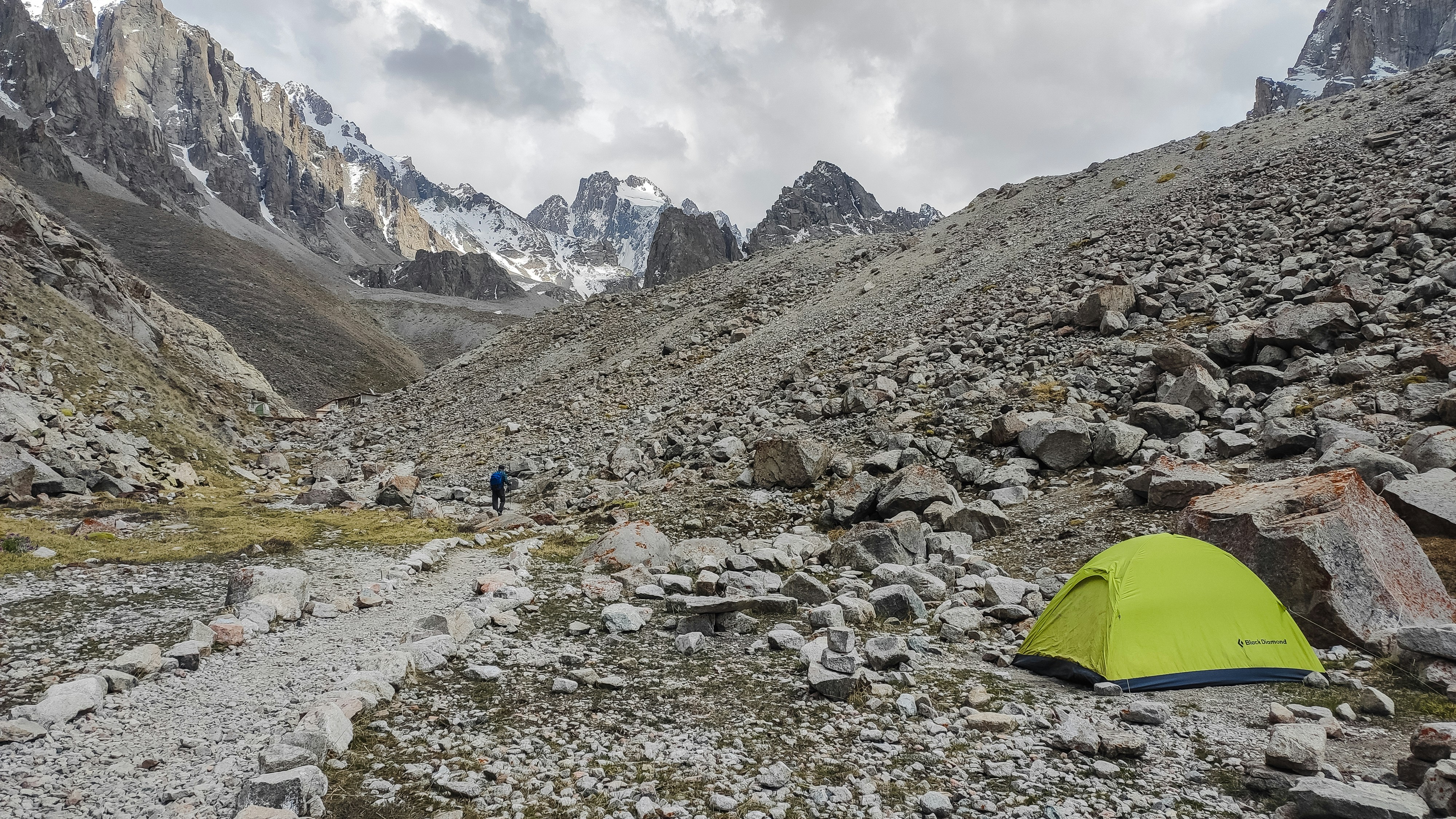 Lime-green tent rests on a rocky talus slope as jagged alpine peaks rise in the background. A lone hiker is visible in the distance.