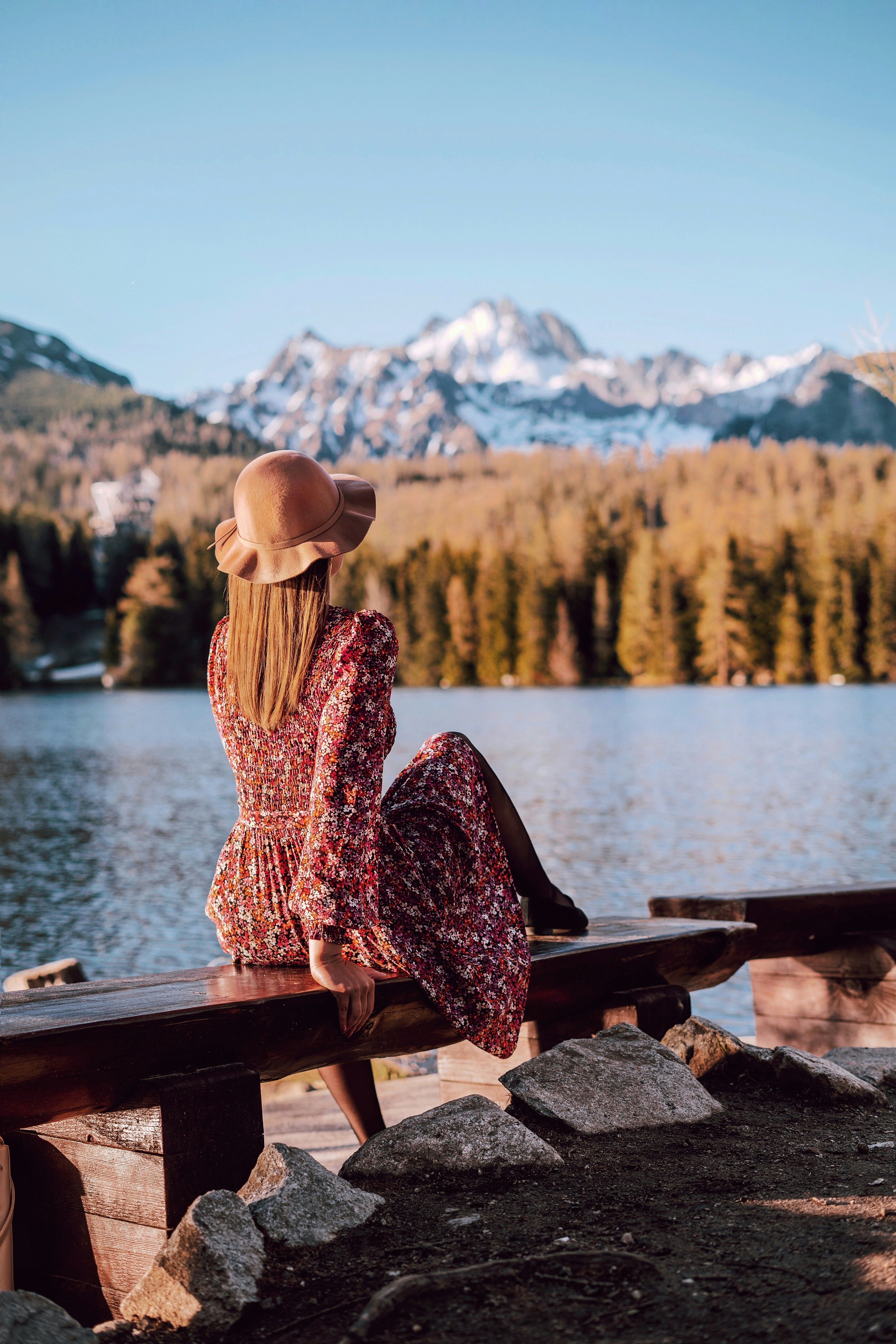Woman in a floral dress and hat sitting on a wooden bench by a serene lake, gazing at snow-capped mountains in the distance.