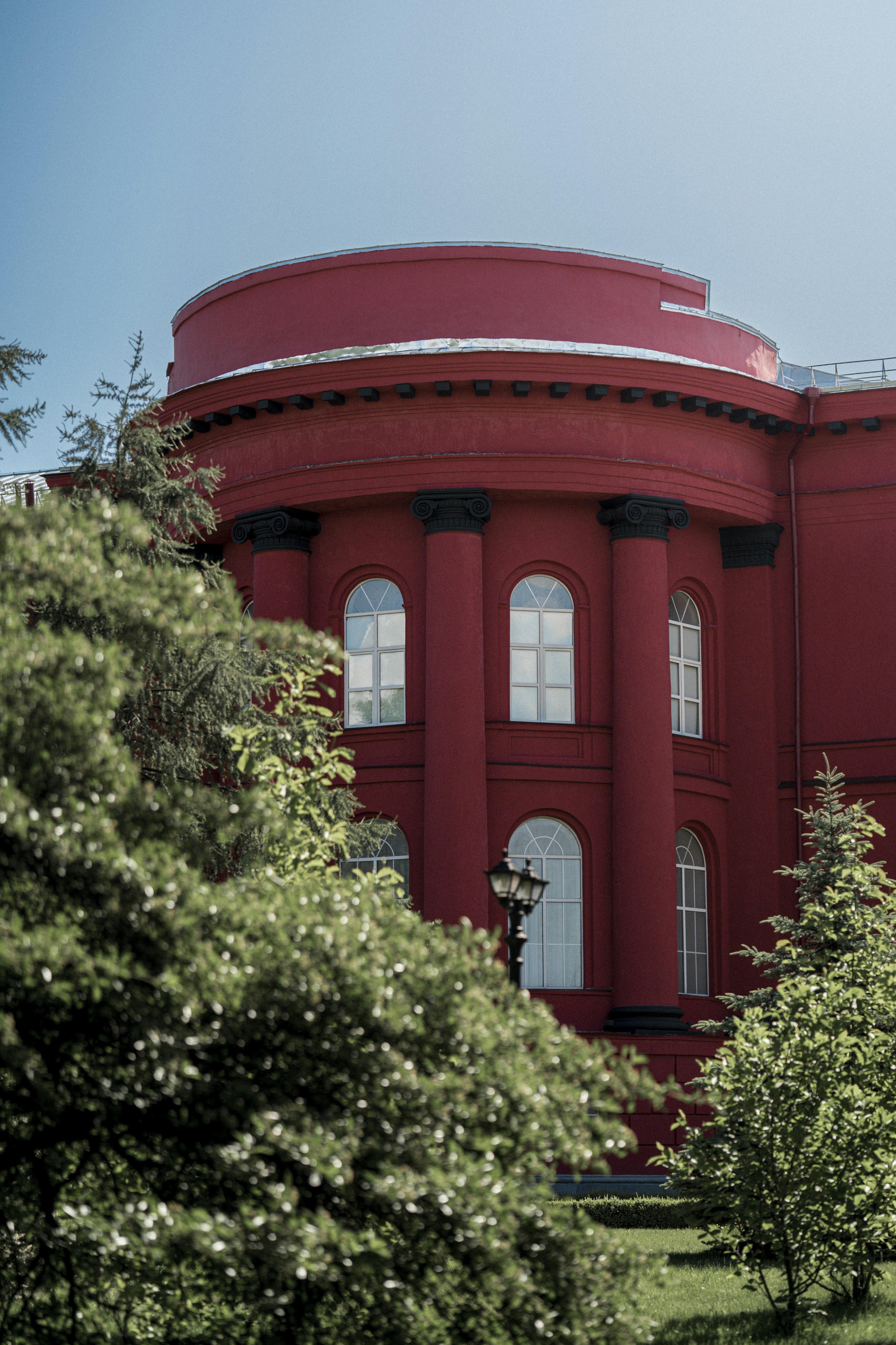 Bold red cylindrical building with arched windows surrounded by lush green trees under a clear blue sky.