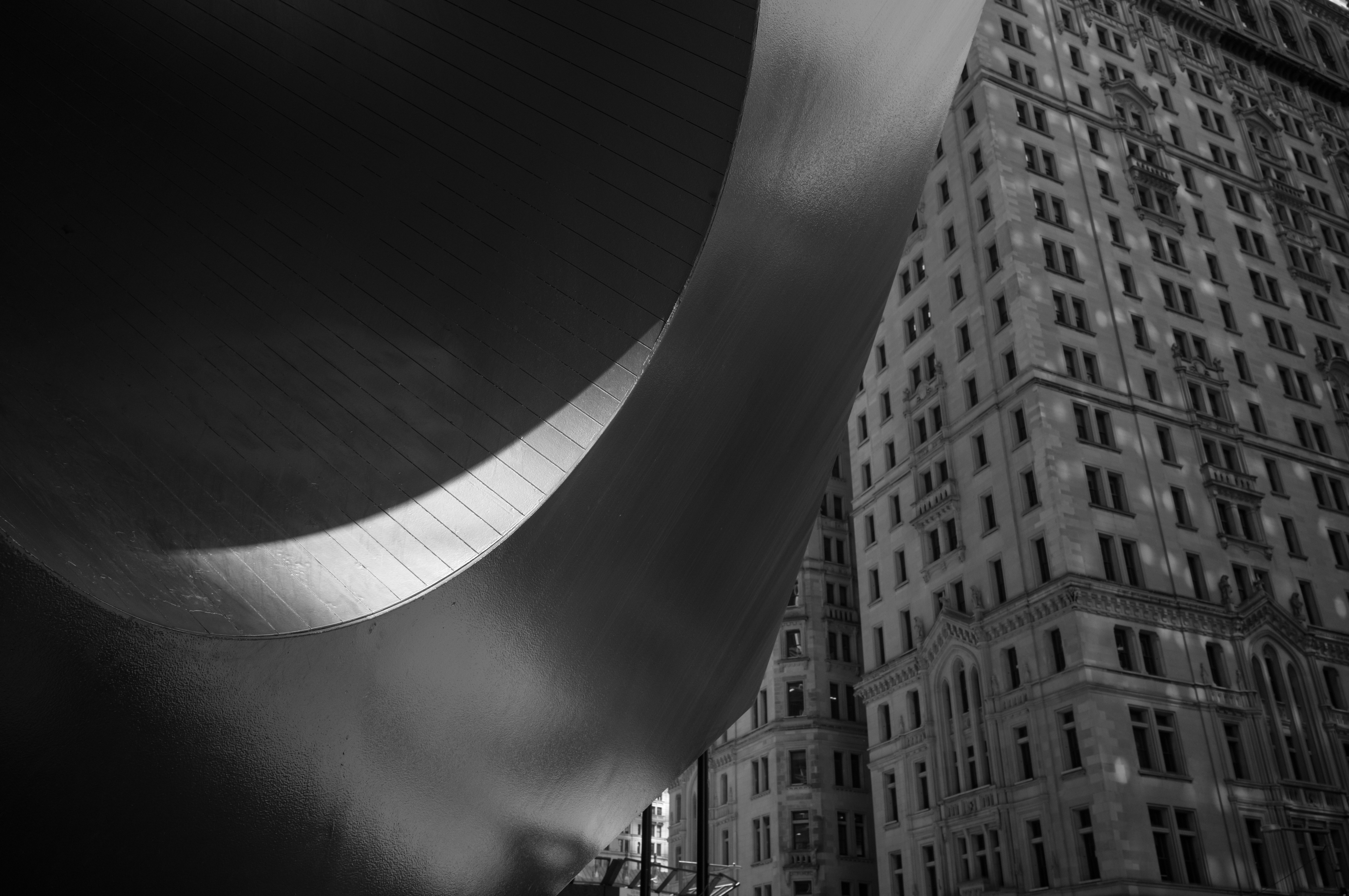High-contrast monochrome photograph of a curved steel ring juxtaposed with a historic brick skyscraper.