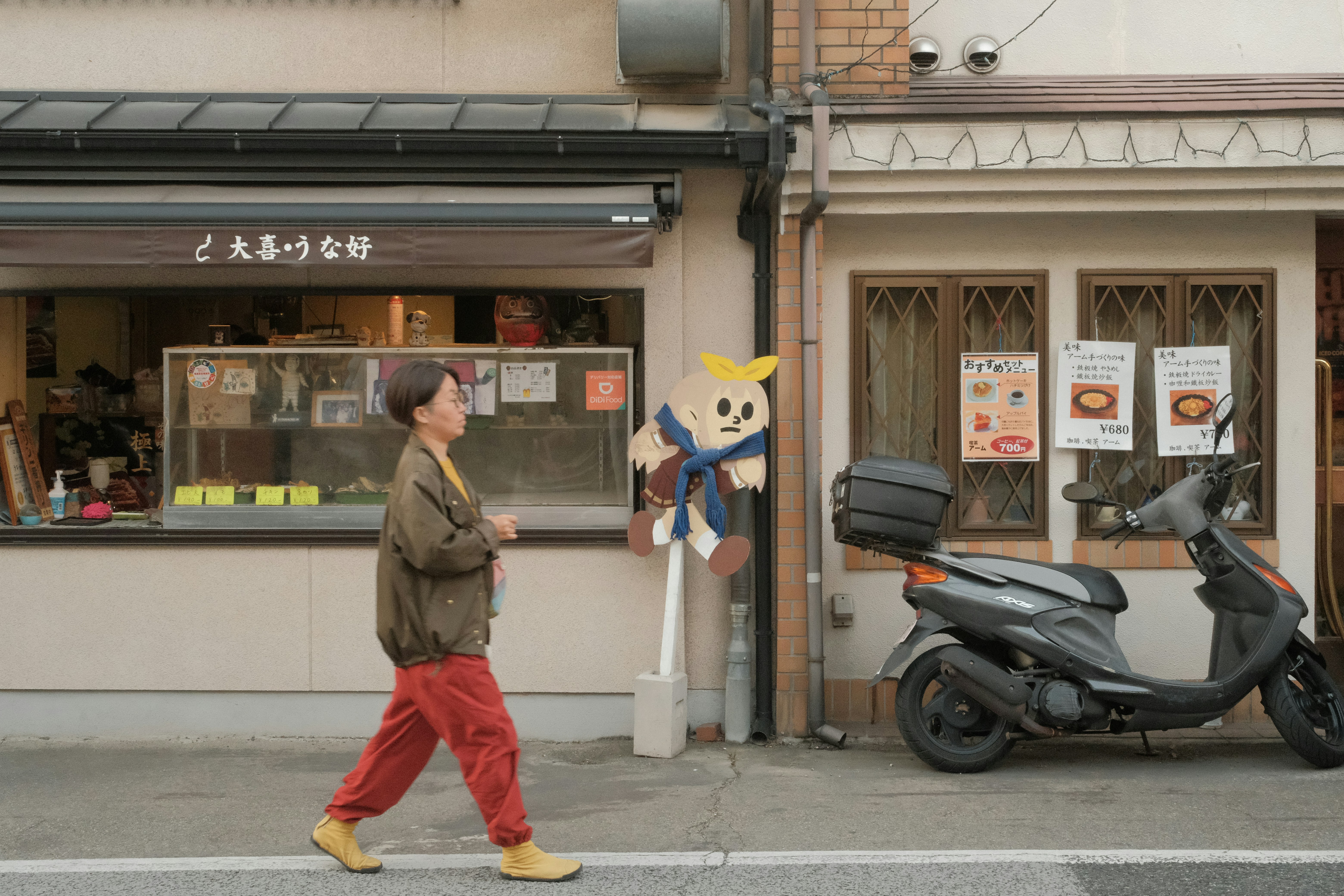A woman strolls past a charming storefront adorned with a playful wooden mascot, capturing the essence of local culture. The scene highlights the vibrant life of the street.