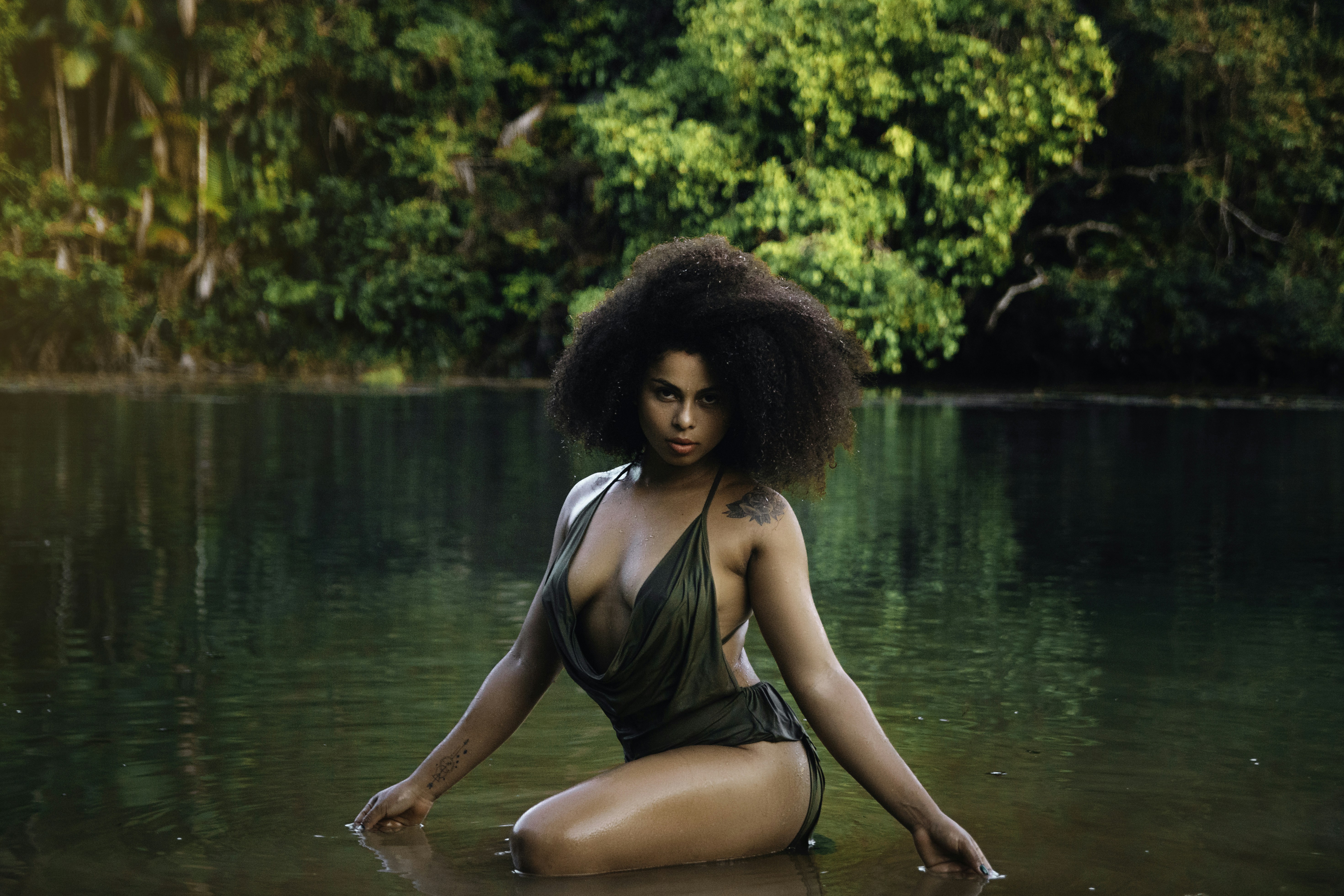 Woman in a green swimsuit kneeling in a river with lush forest backdrop.