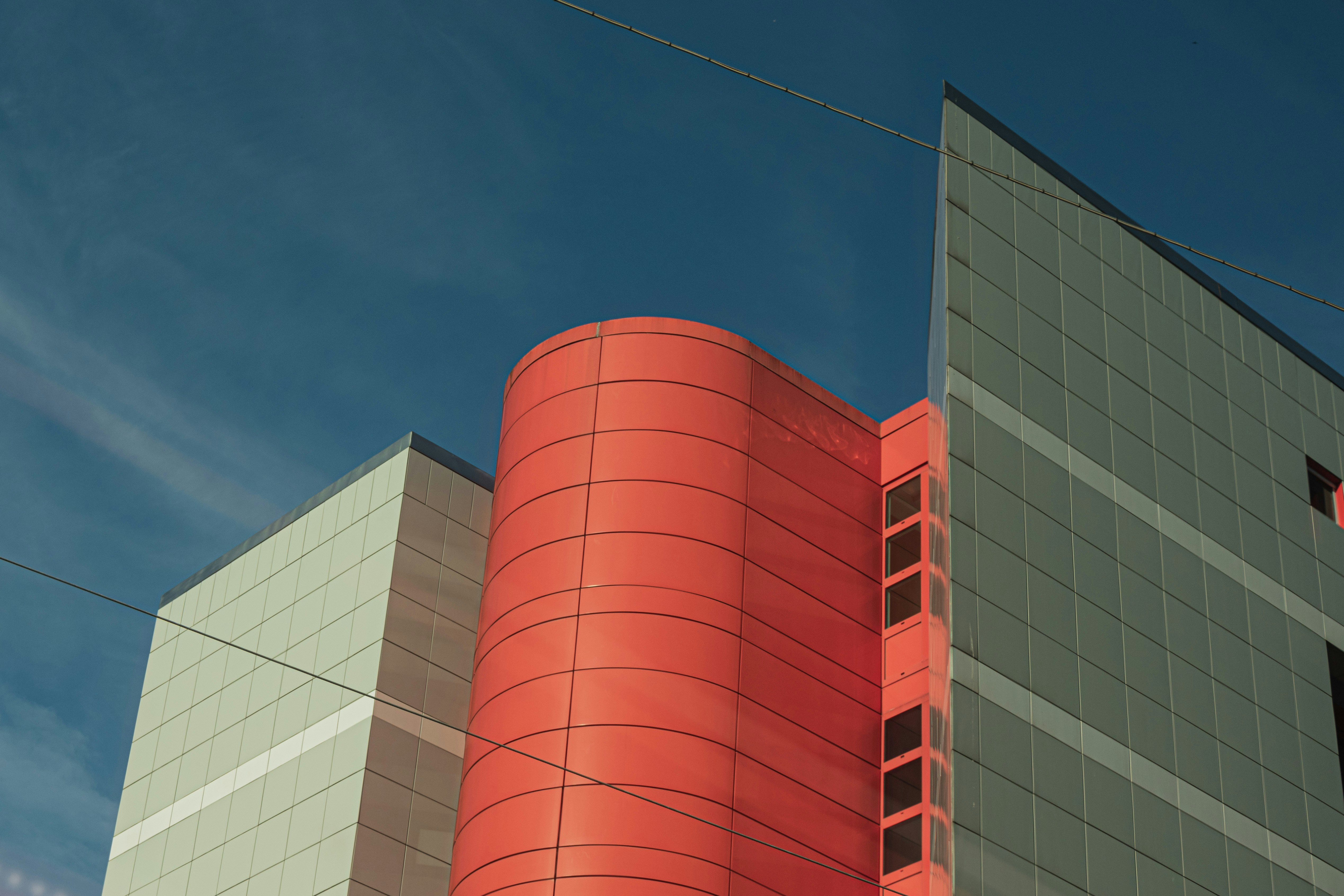 Modern building facade featuring a striking red cylindrical element alongside angular structures under a clear blue sky.