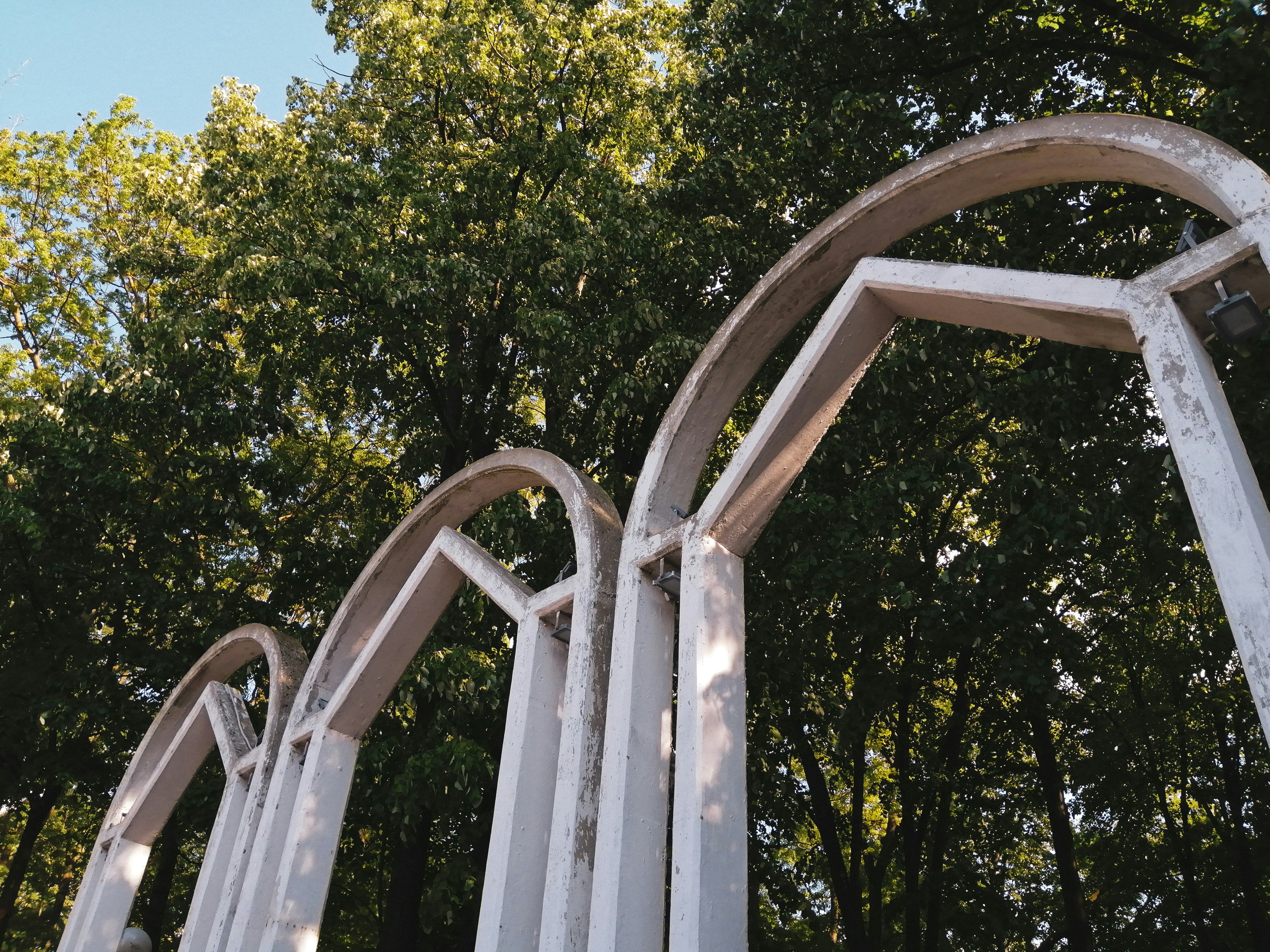 White metal arches create a repeating corridor through a sunlit park, with green treetops forming a leafy backdrop.