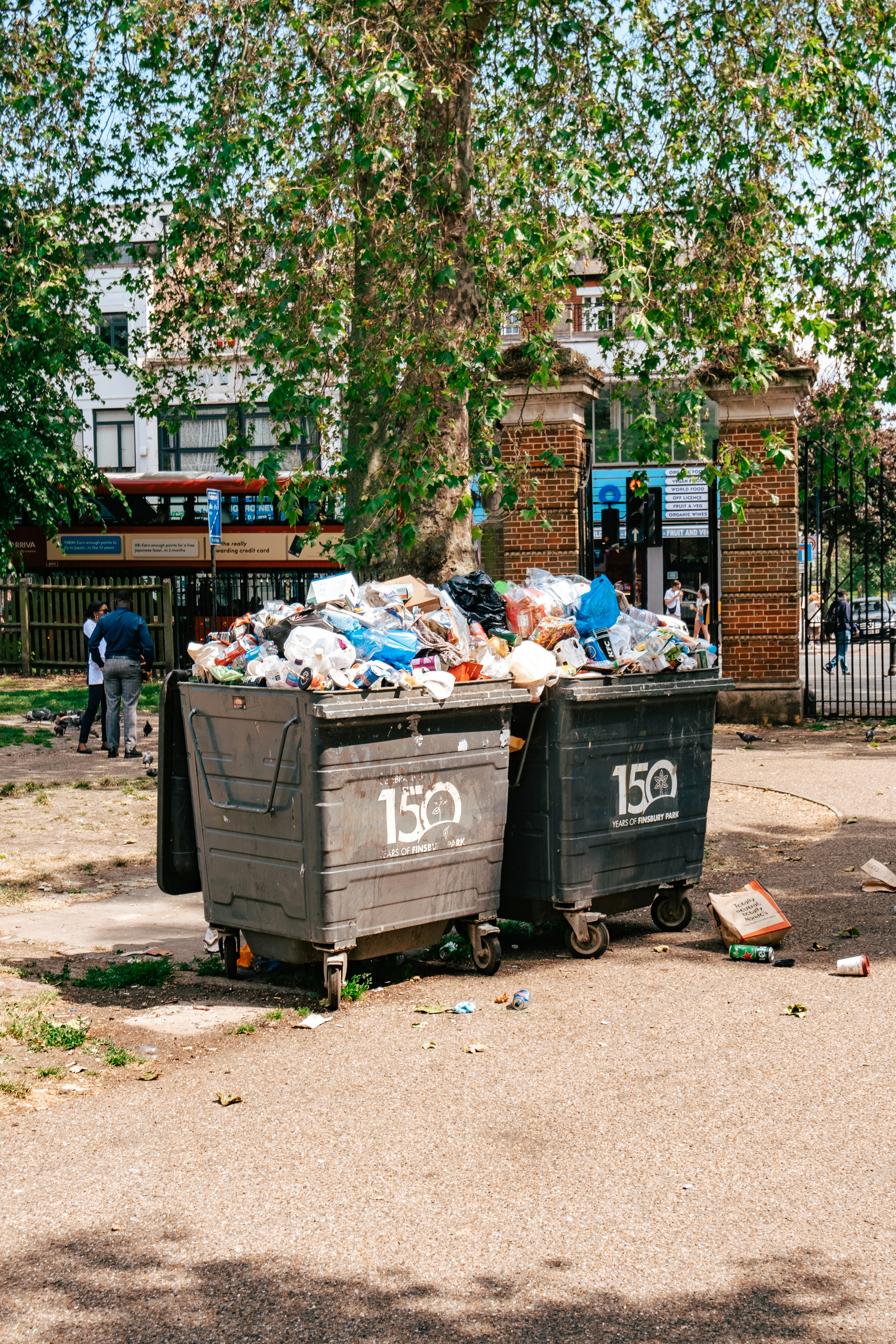 Overflowing trash bins in a city park highlight the challenges of urban waste disposal. Lush green foliage frames the scene, contrasting with the clutter of discarded items.