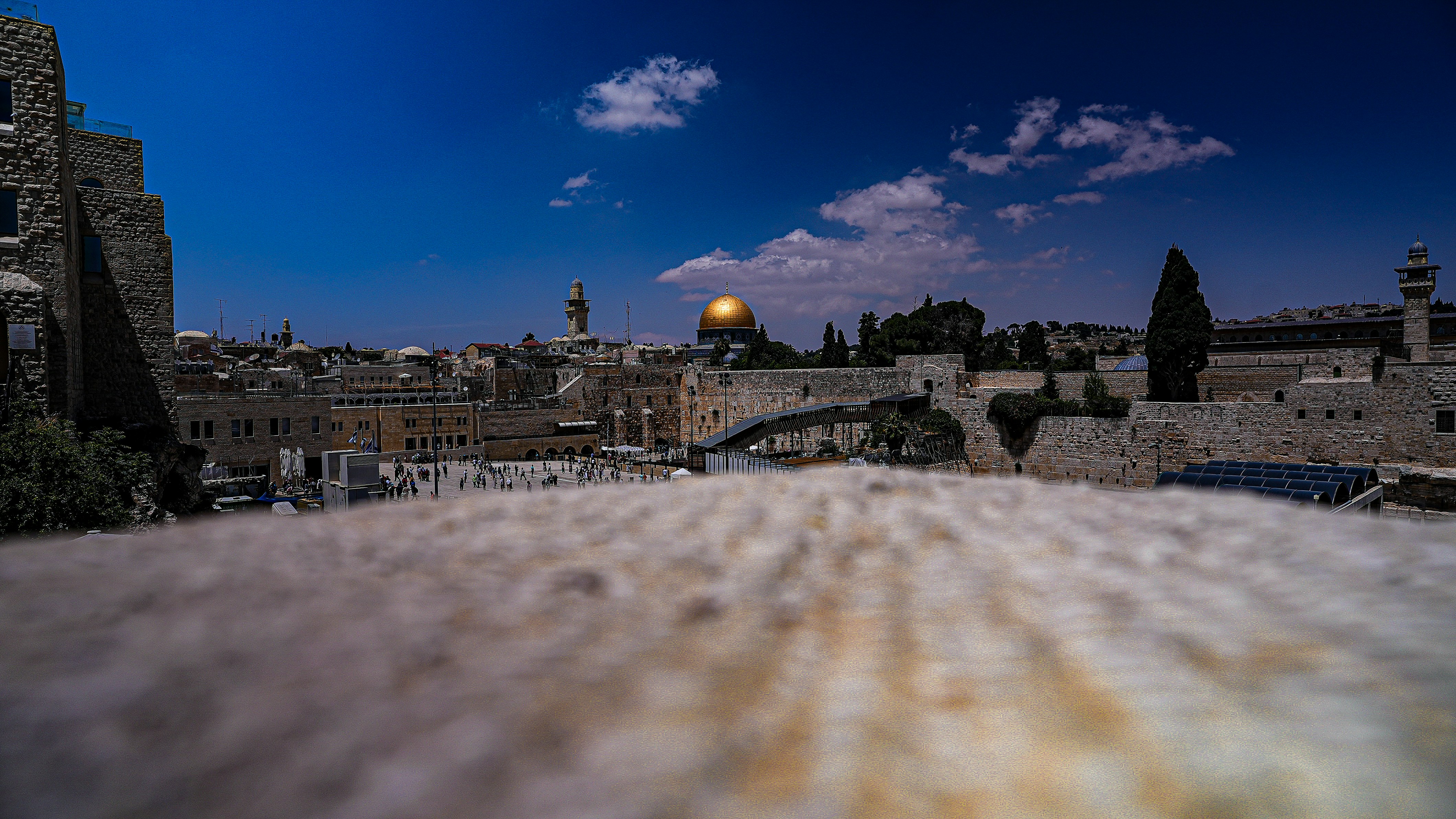Golden Dome of the Rock rises above ancient Jerusalem walls under a bright blue sky. Foreground stone railing is blurred, framing a historic cityscape.
